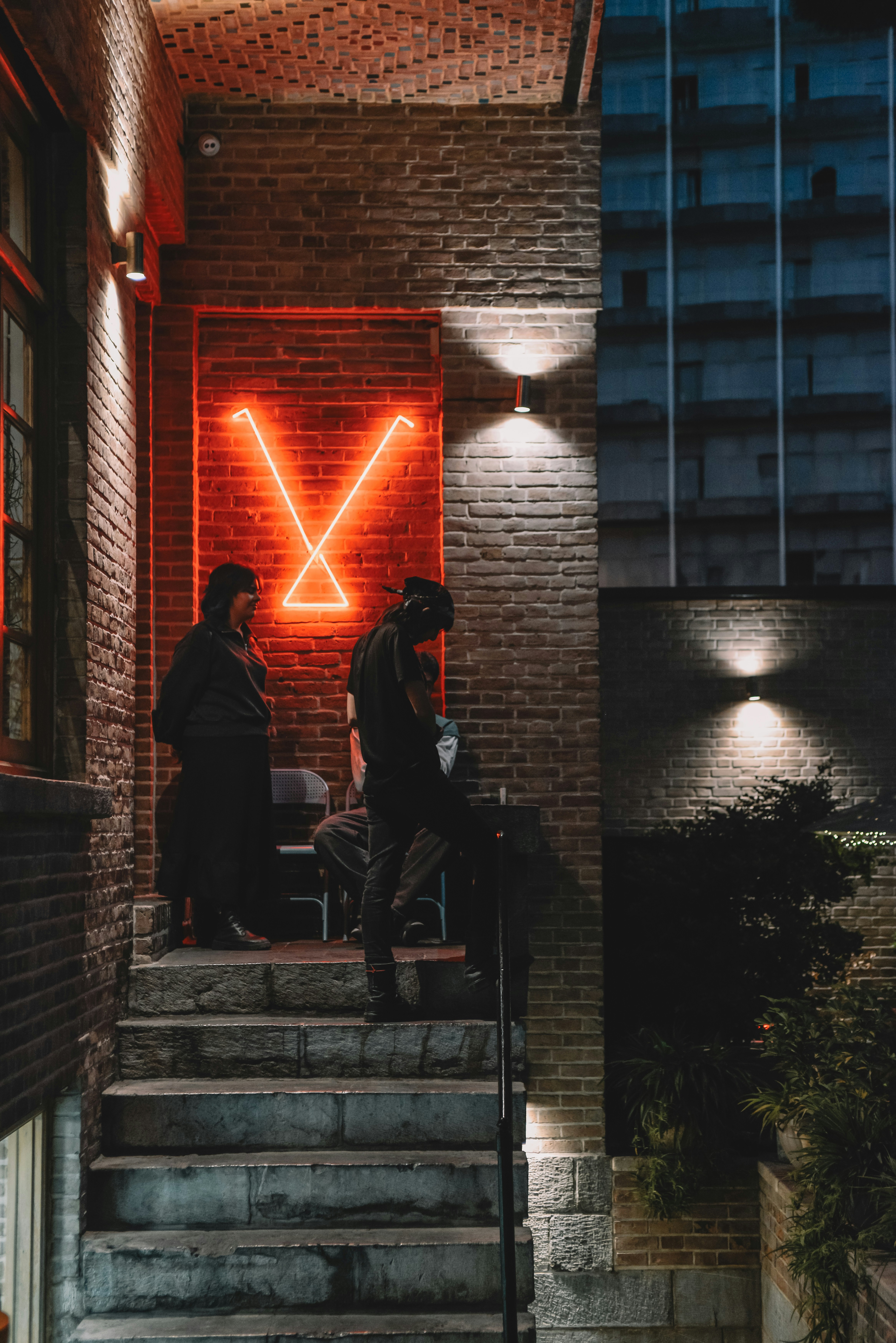 Two figures stand on stone steps illuminated by a vibrant red neon sign, contrasting with the cool tones of the surrounding architecture.