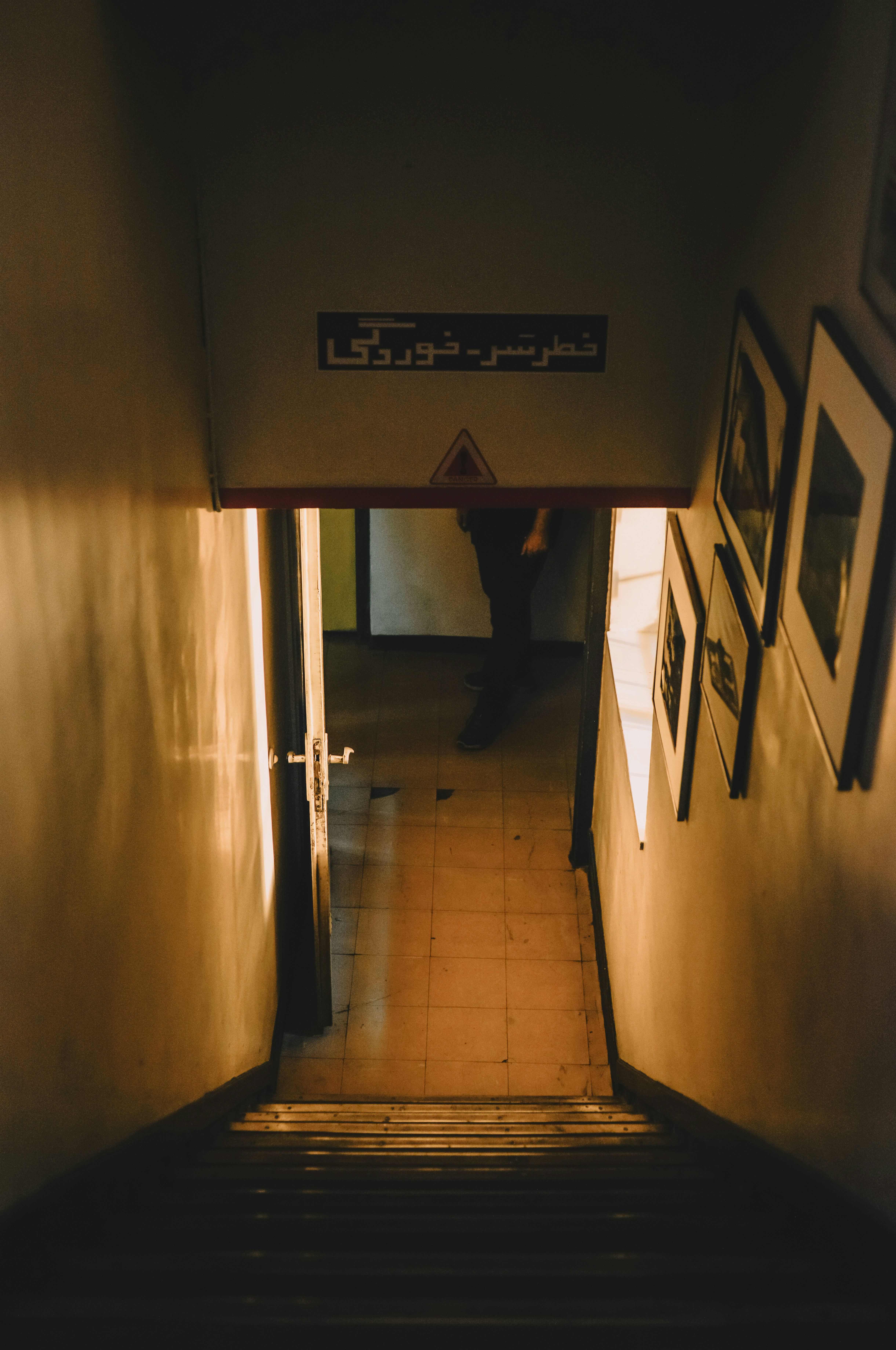 Dimly lit staircase leading down to an open door, with a warning sign above and framed images along the wall.