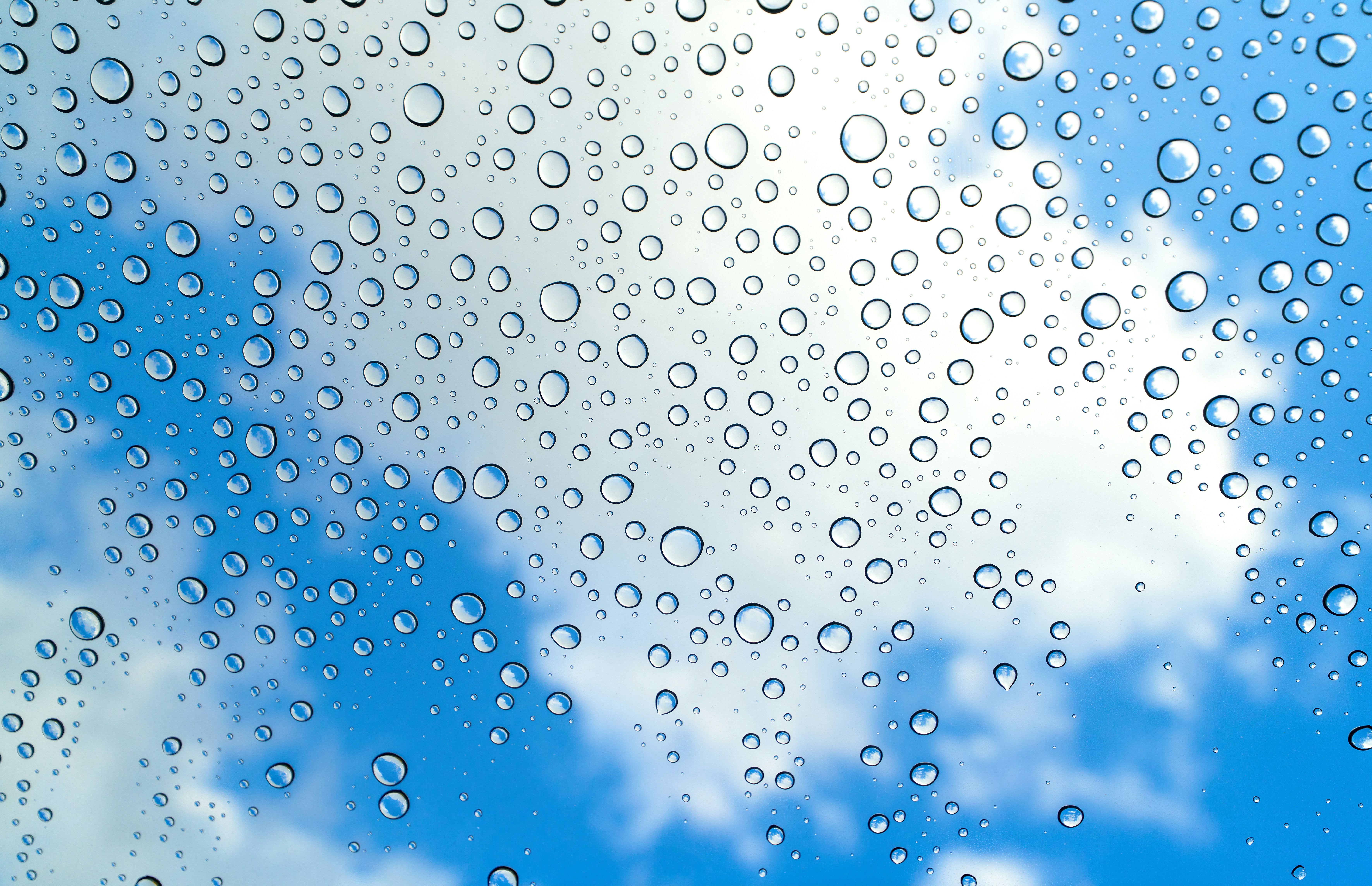 Raindrops clinging to a glass surface, creating a textured view of a blue sky dotted with fluffy clouds.
