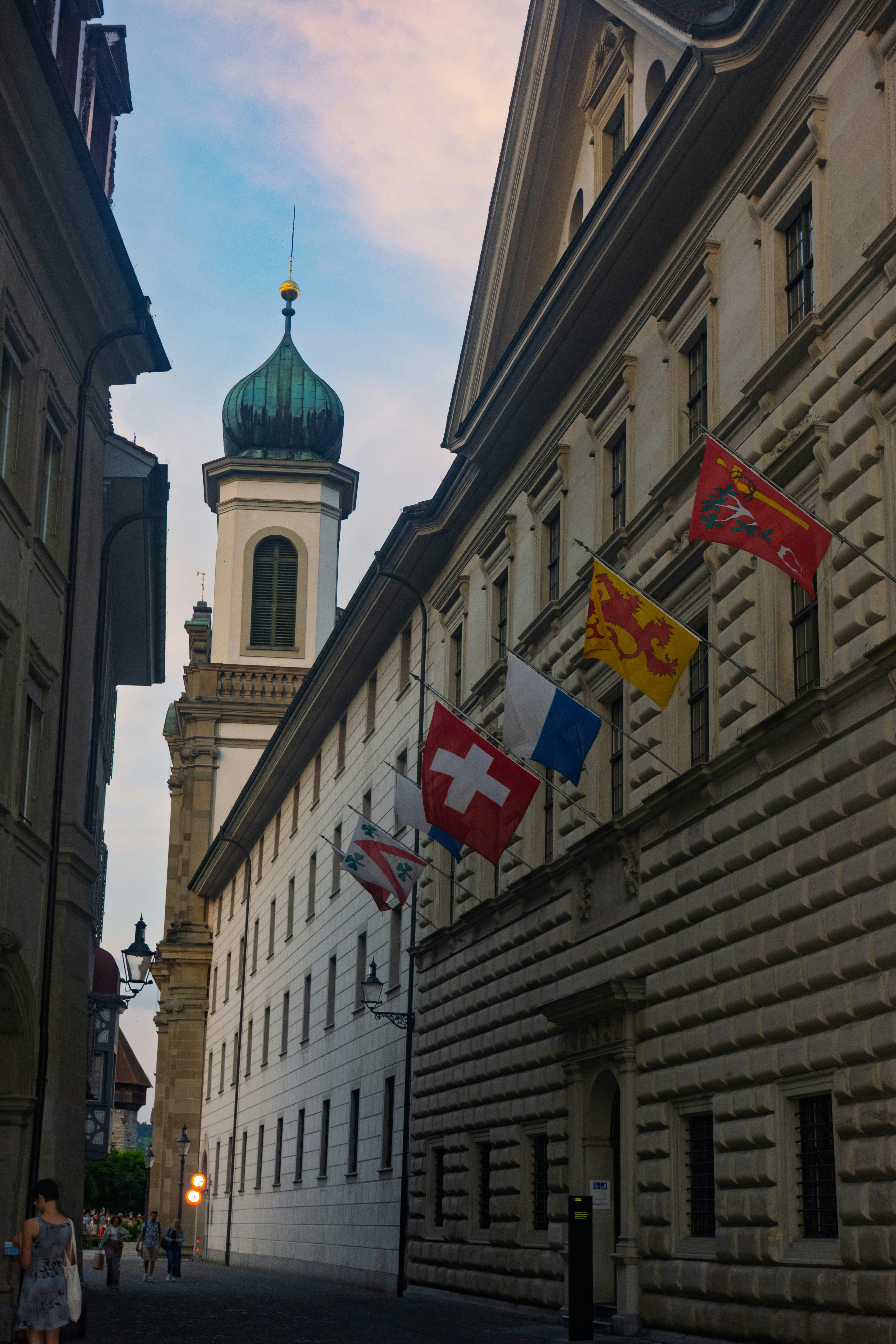 Historic building with colorful flags lining the walls, framed by narrow cobblestone street and a towering clock tower in the background.