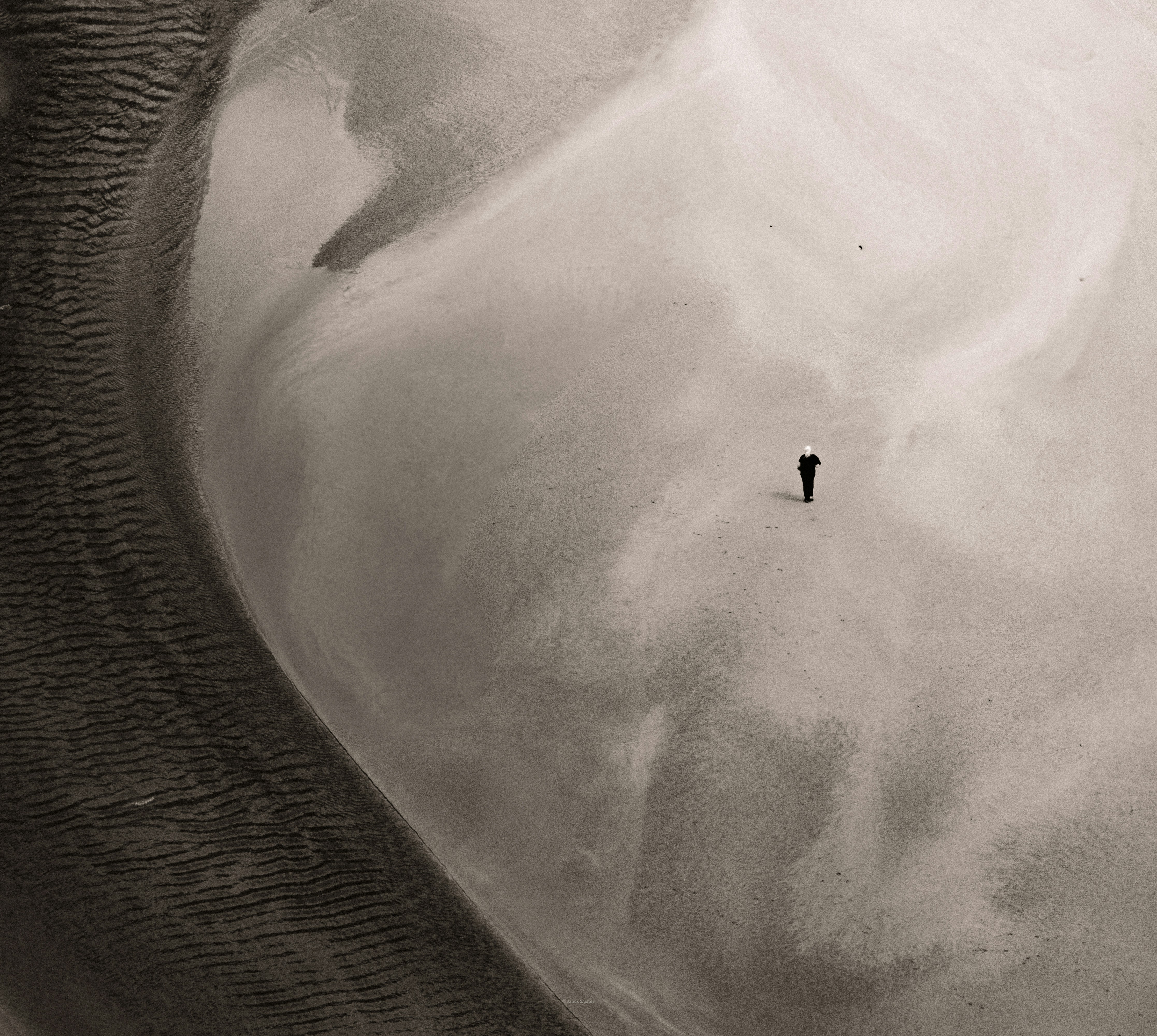 A lone figure, walking across a vast, sand-like terrain. The minimalist composition, highlighting the emptiness of the landscape, evokes a sense of solitude and reflection.