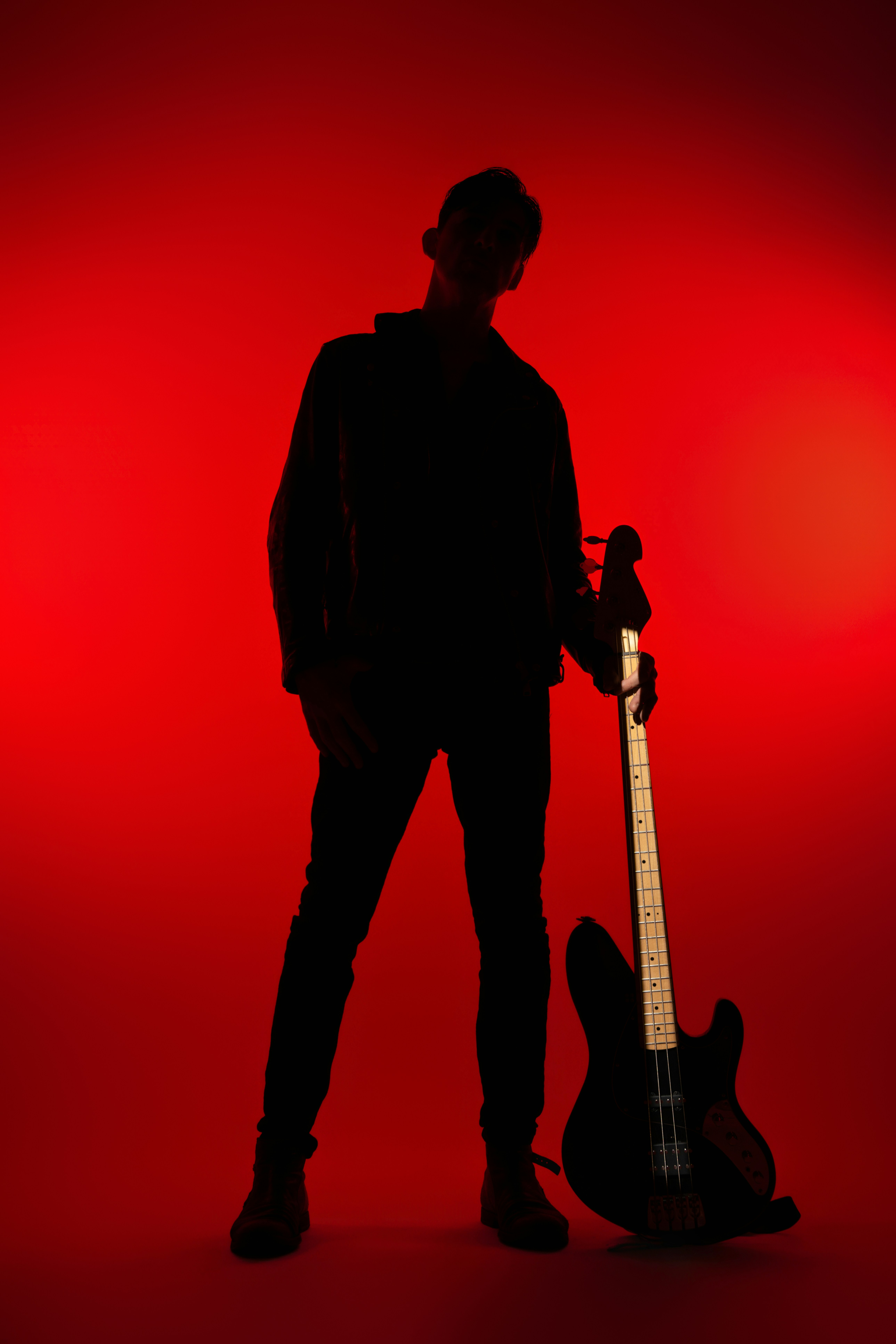 Silhouette of a musician standing against a vibrant red background, holding a bass guitar. The stark contrast highlights the figure's pose and the instrument's shape.