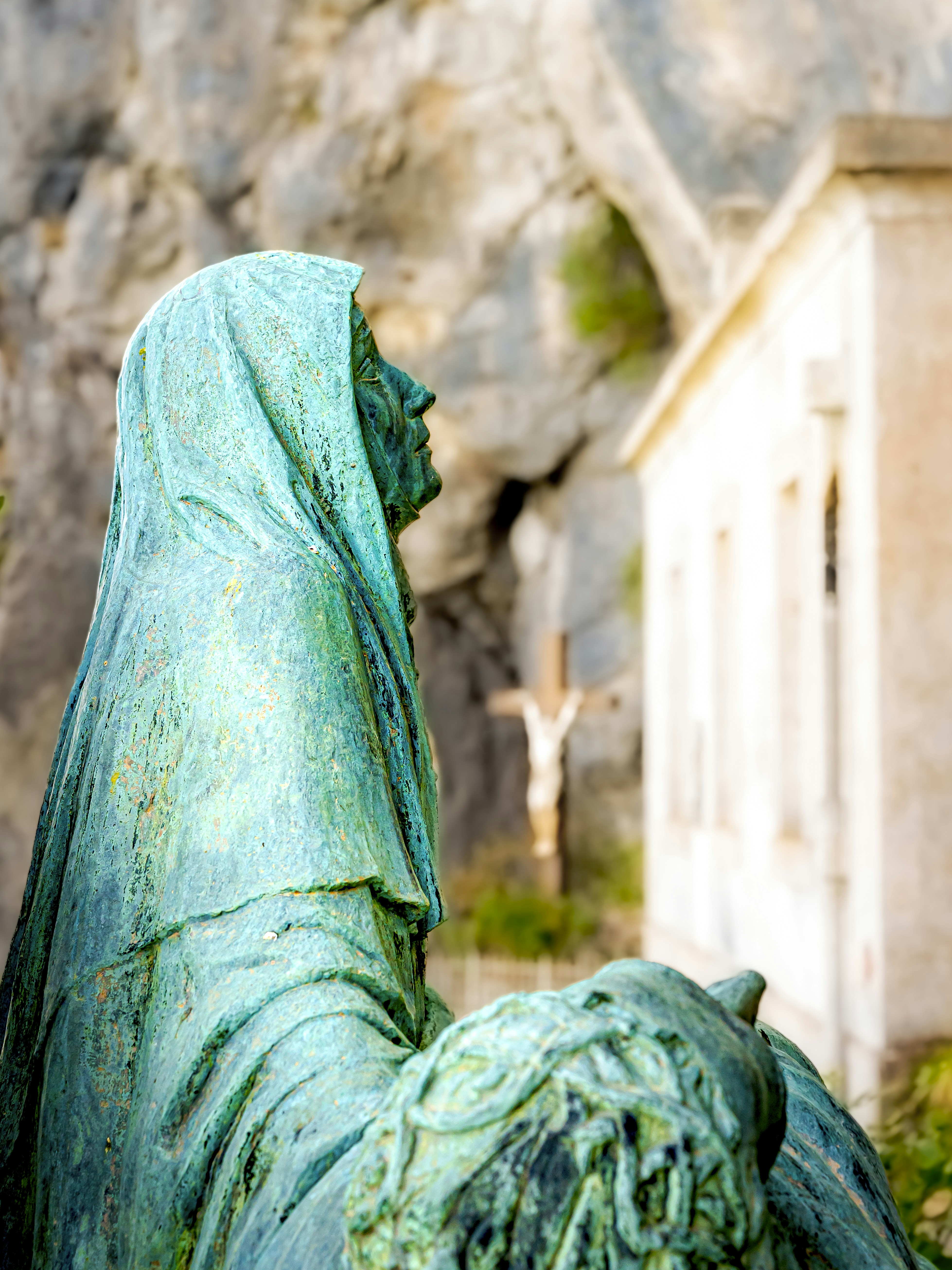 A weathered bronze statue of a cloaked figure gazes contemplatively toward a distant cross, set against a backdrop of rugged stone and soft greenery.