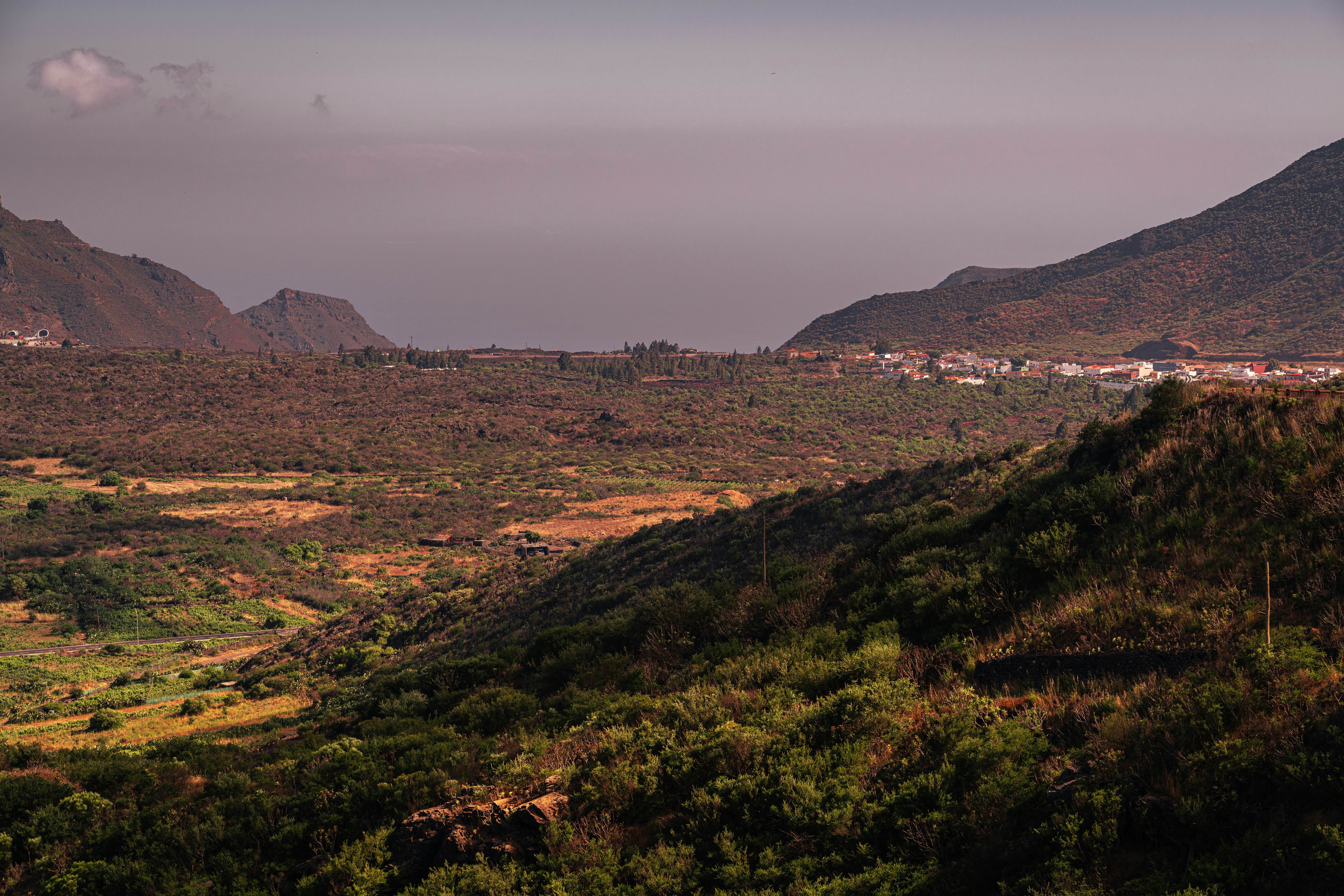 Lush green hills cradle a tranquil valley, dotted with distant settlements and framed by rugged mountains under a soft sky.