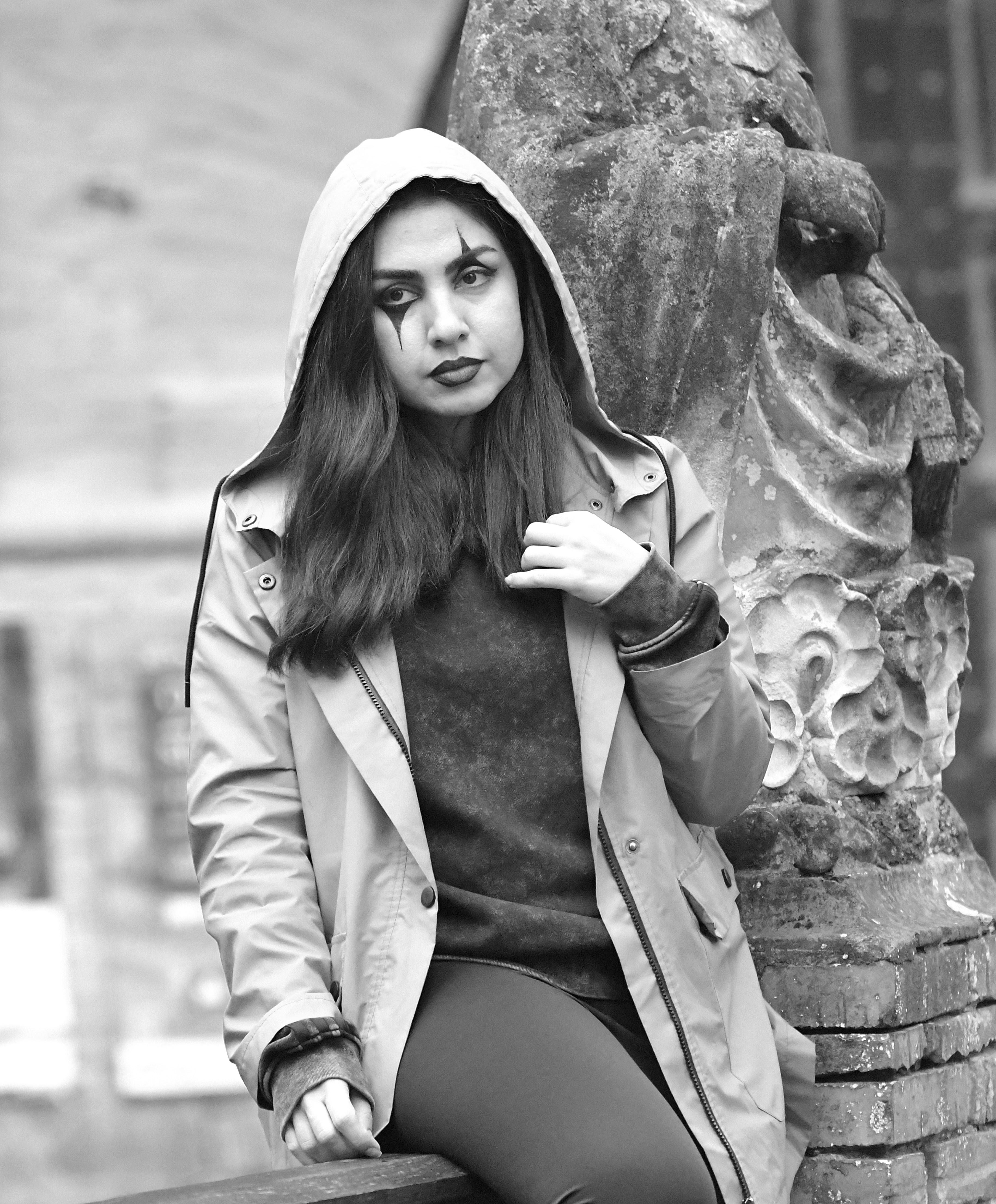 A young woman in a hooded coat leans against a weathered statue, exuding a sense of introspection and mystery. The monochromatic tones enhance the emotional depth of the scene.