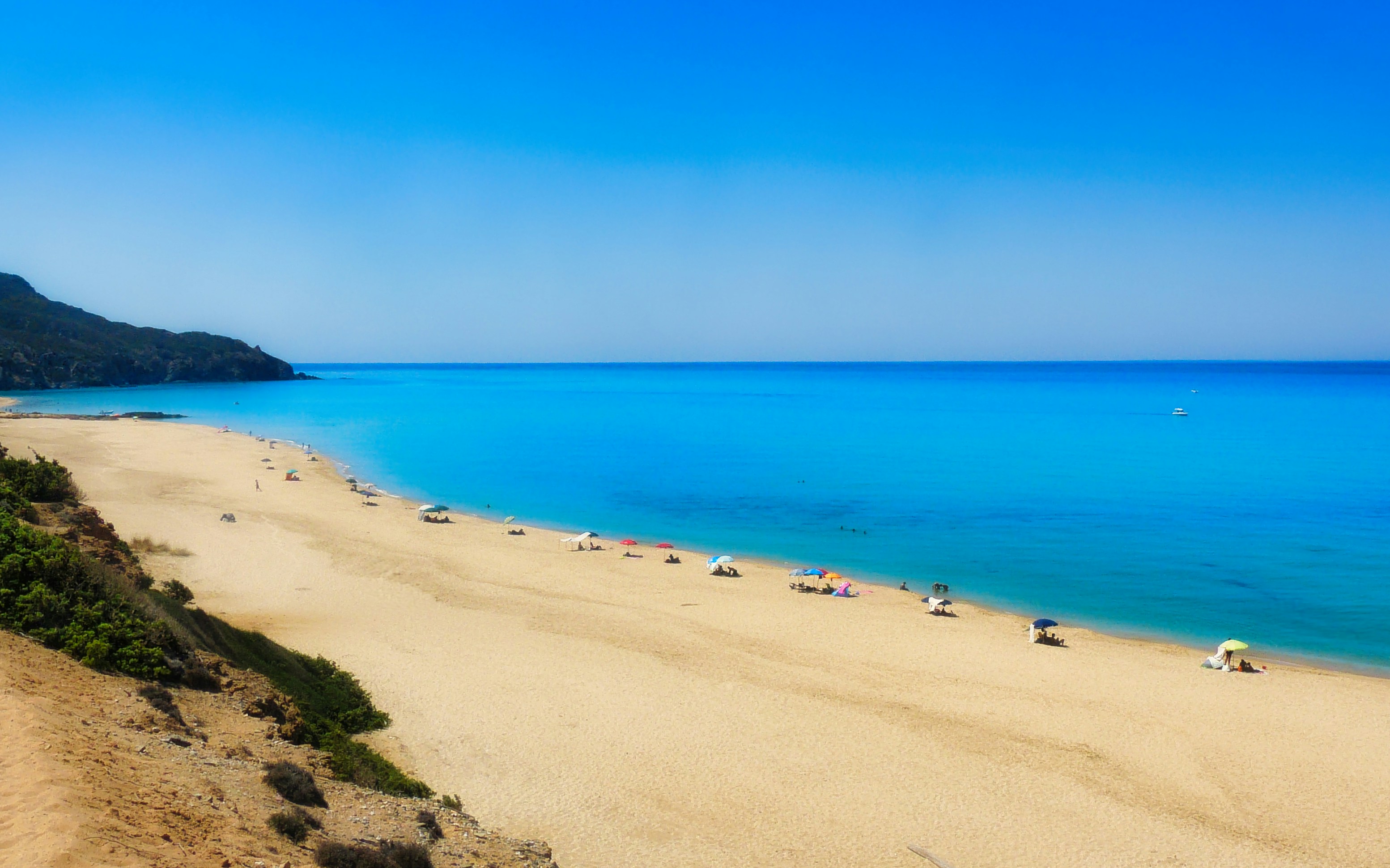 Expansive sandy beach with sunbathers and colorful umbrellas under a clear blue sky. The tranquil sea stretches into the horizon.