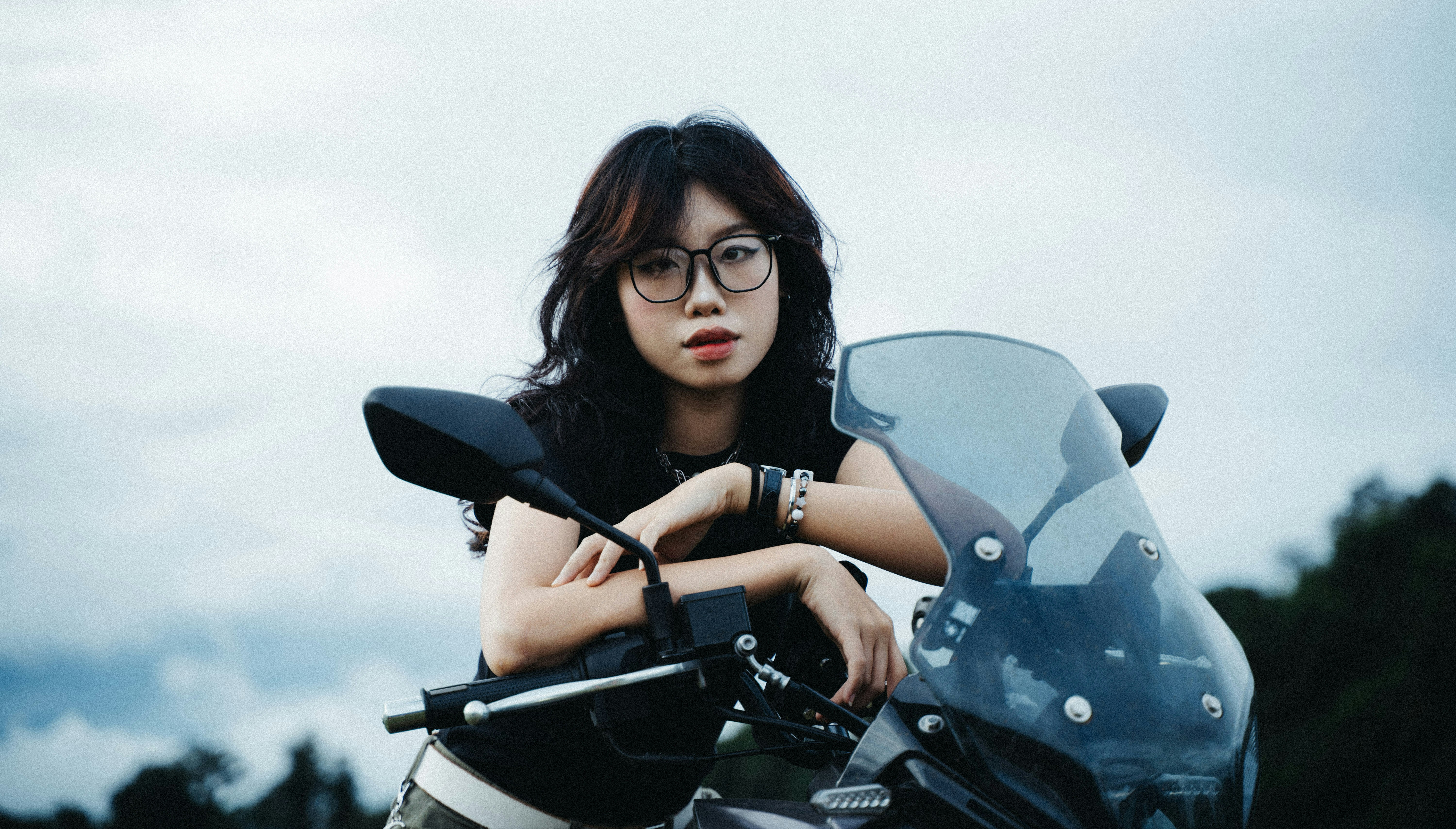 Young woman with glasses resting on a motorcycle, exuding a calm demeanor against a backdrop of dramatic clouds.