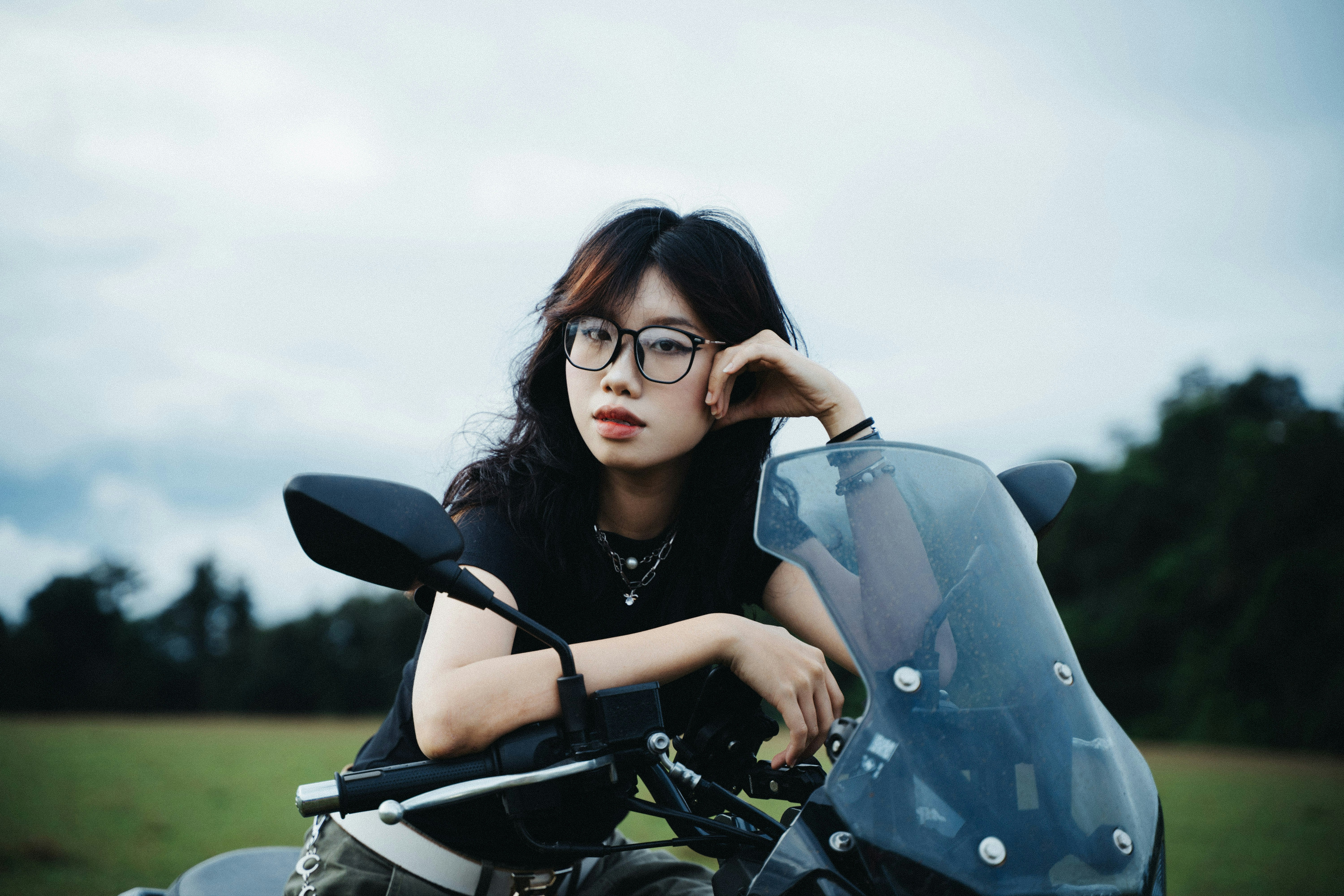 Young woman with glasses leaning on a motorcycle in an open field under a cloudy sky.