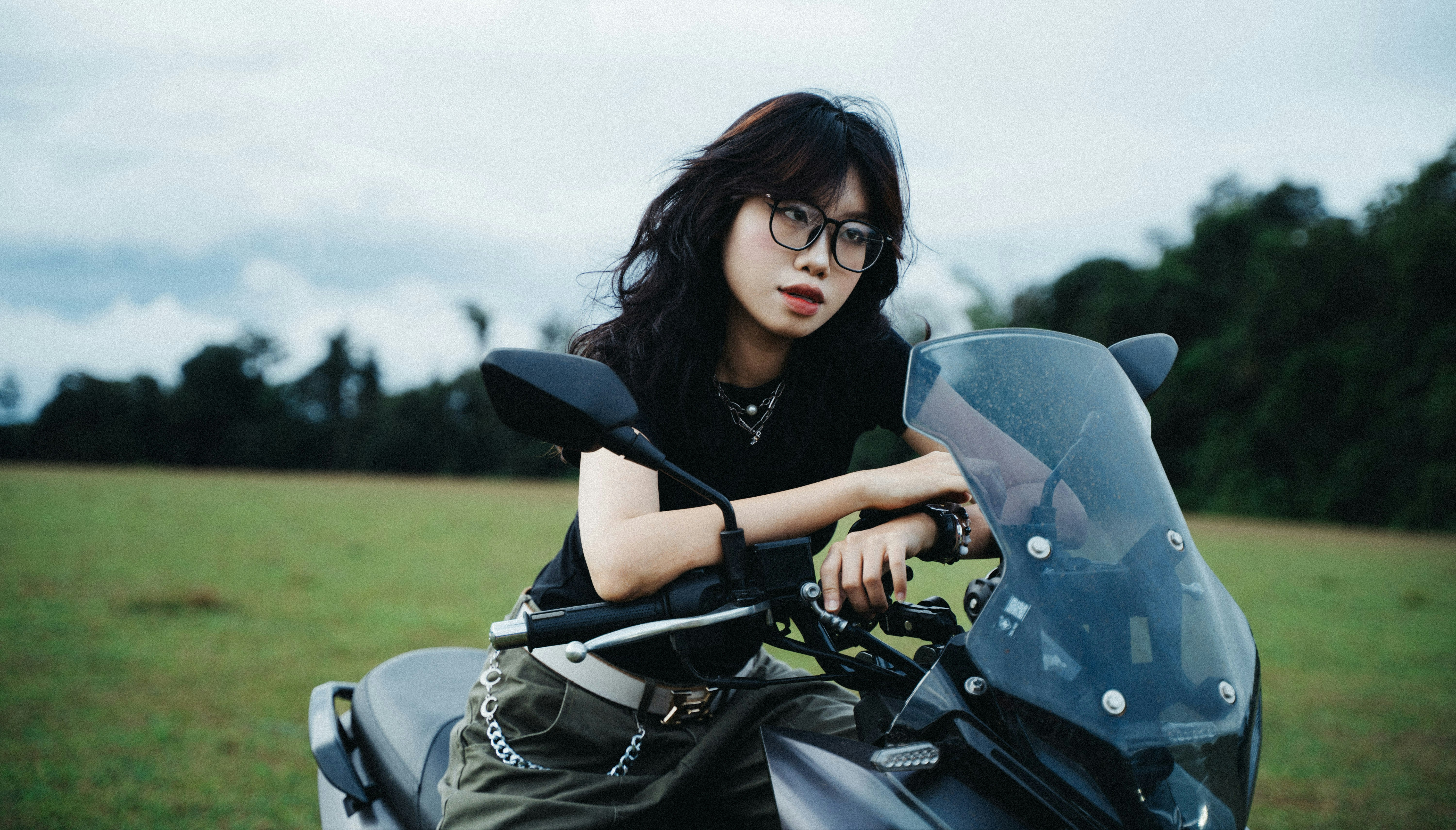 Young woman with glasses perched on a motorcycle, surrounded by a lush green landscape under a cloudy sky.