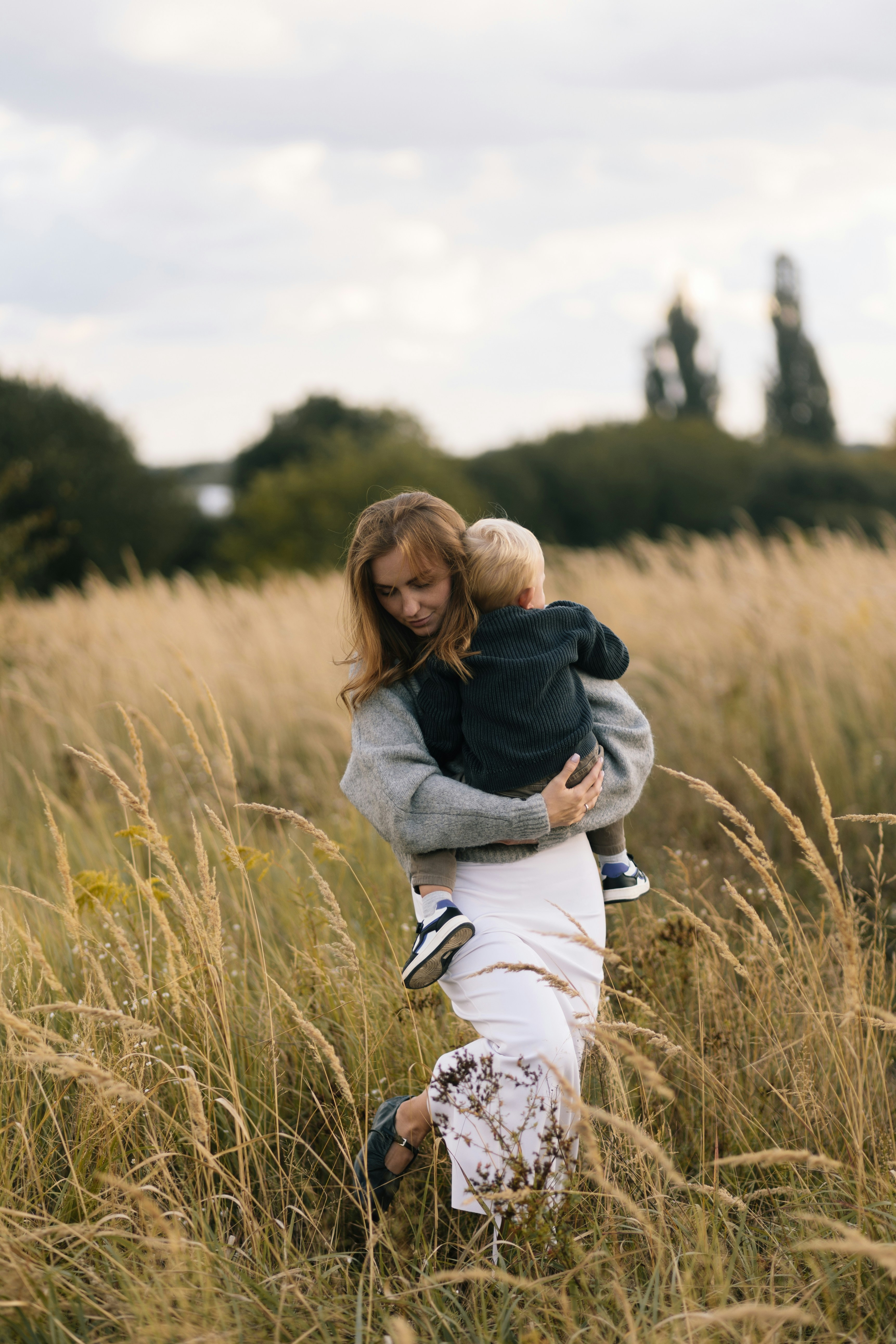 Woman holding a child in a field of tall grass, surrounded by a soft, natural landscape. The scene conveys warmth and connection.