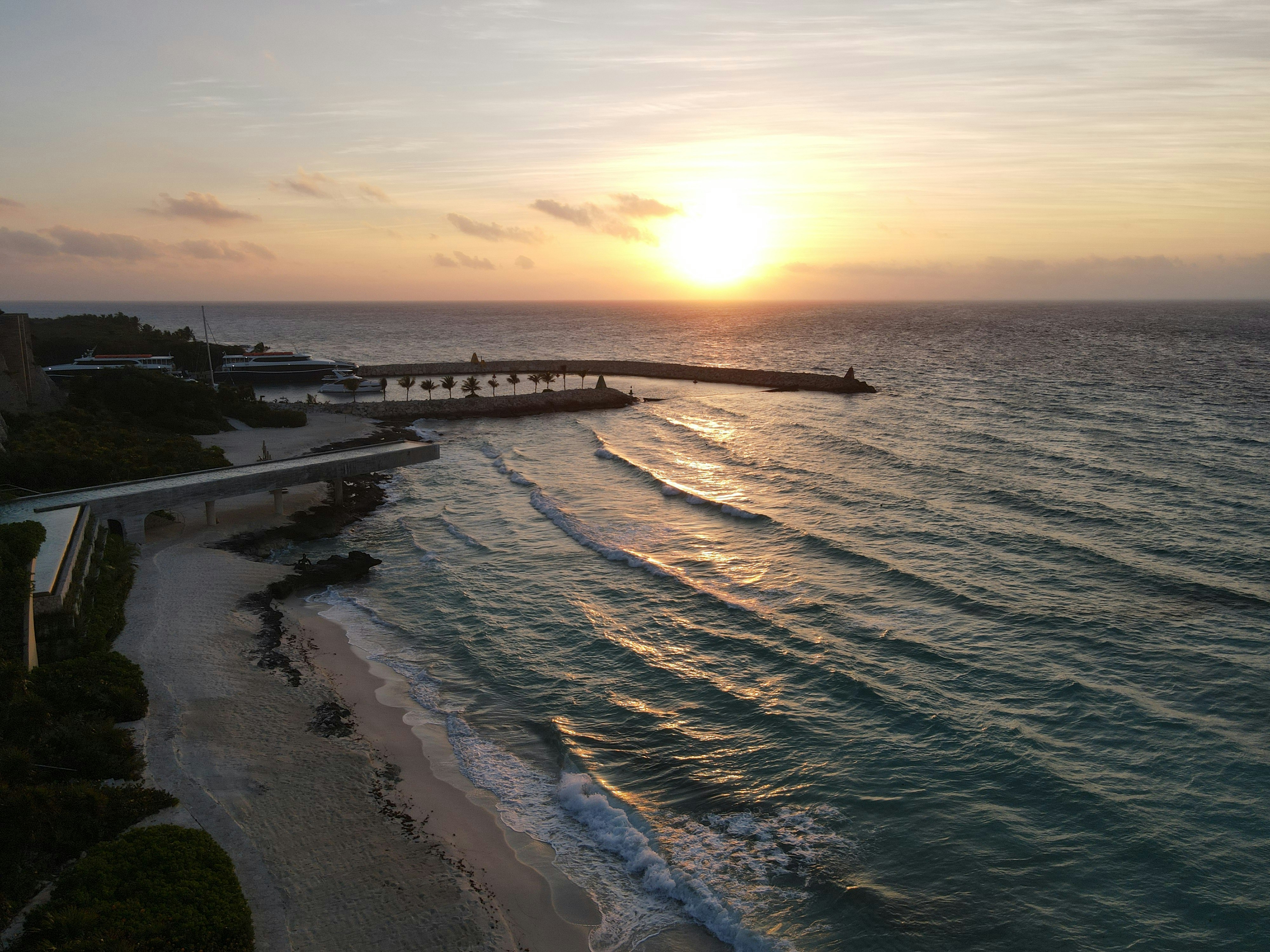 Golden sun dipping below the horizon, casting reflections on gentle ocean waves near a coastal promenade.