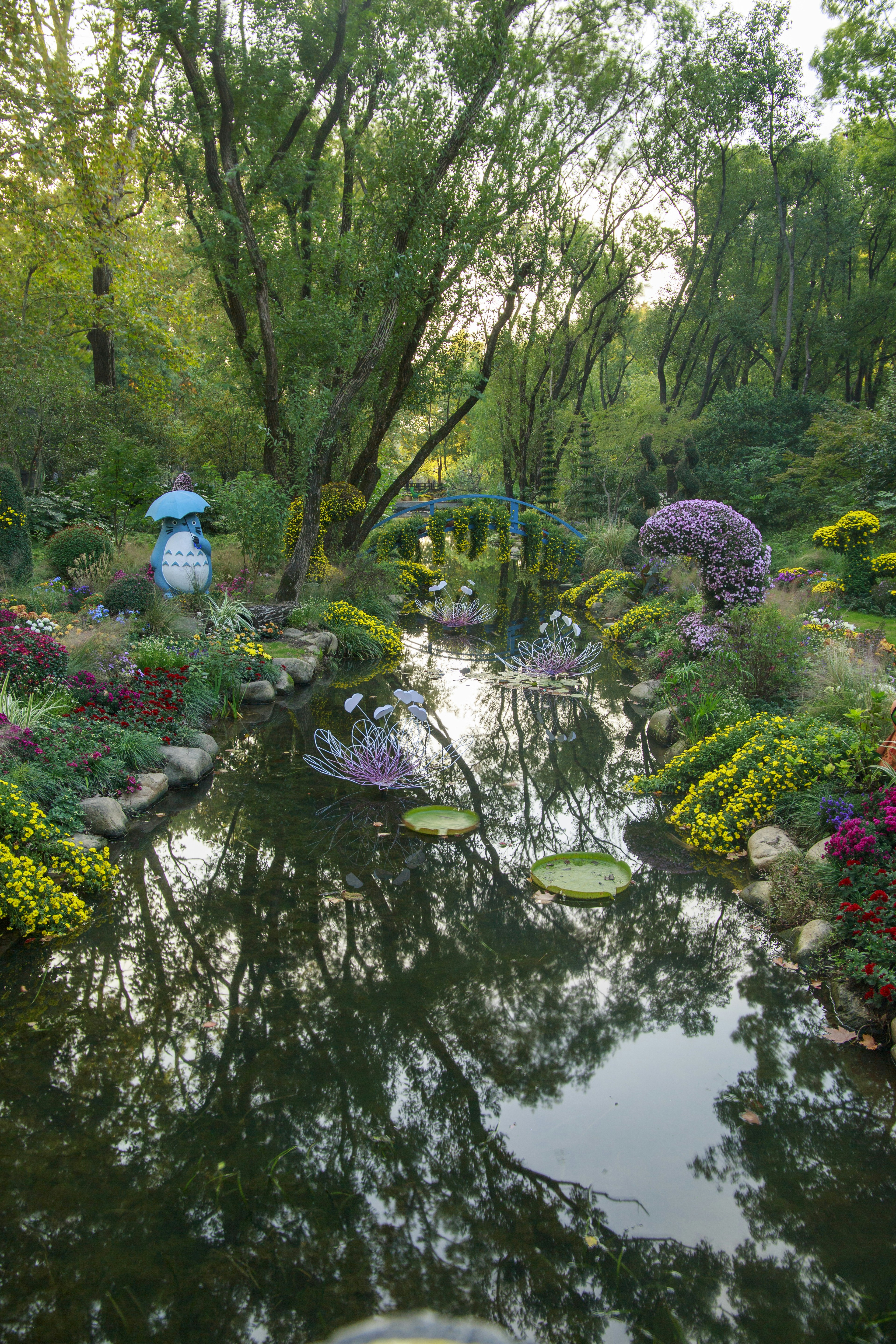 A serene garden scene featuring colorful flowers and whimsical decorations, with reflections shimmering in the tranquil pond. The lush greenery envelops the area, creating a peaceful atmosphere.