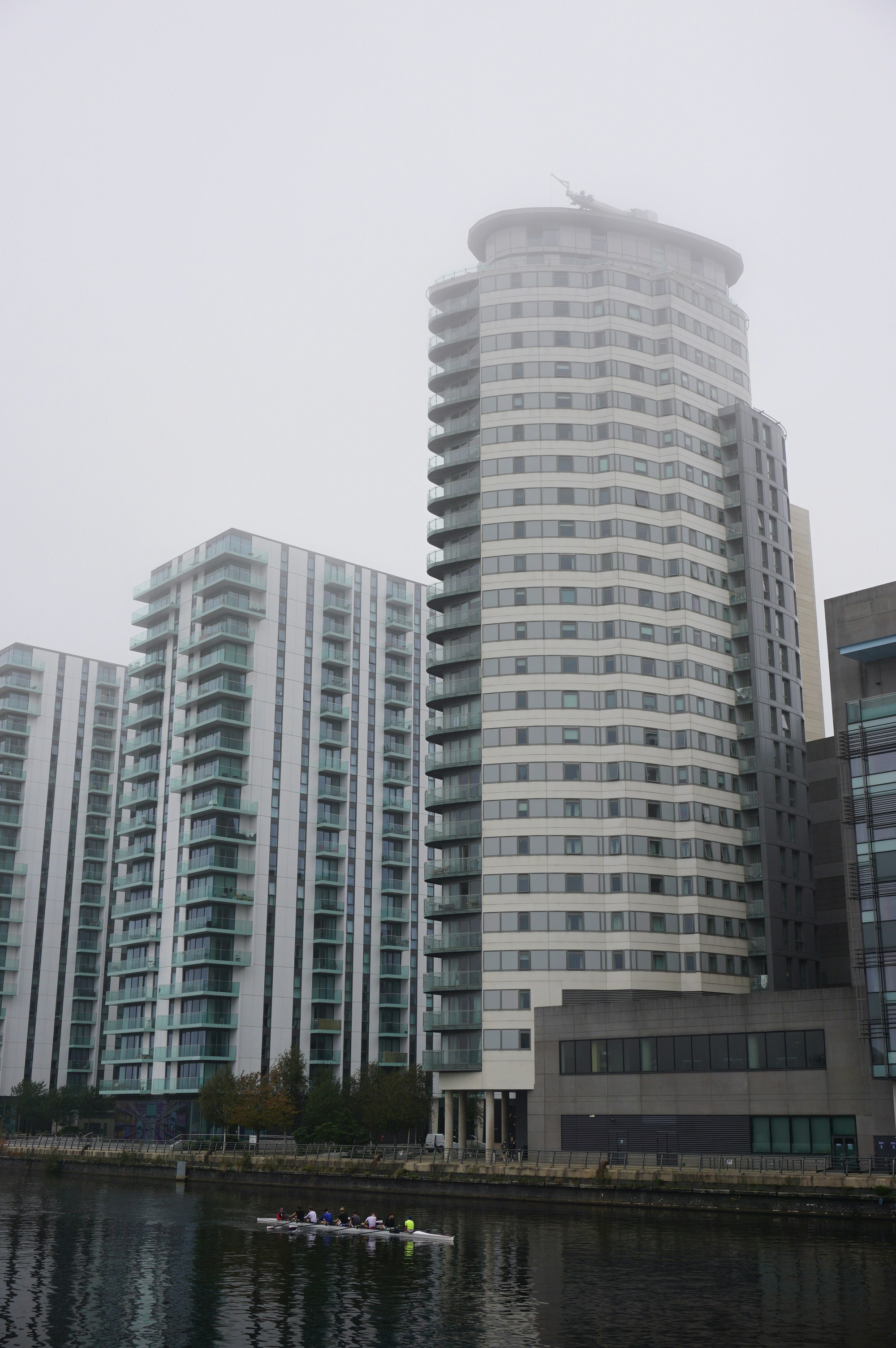 Rowers glide smoothly along a misty waterway, framed by modern high-rise buildings disappearing into the fog.