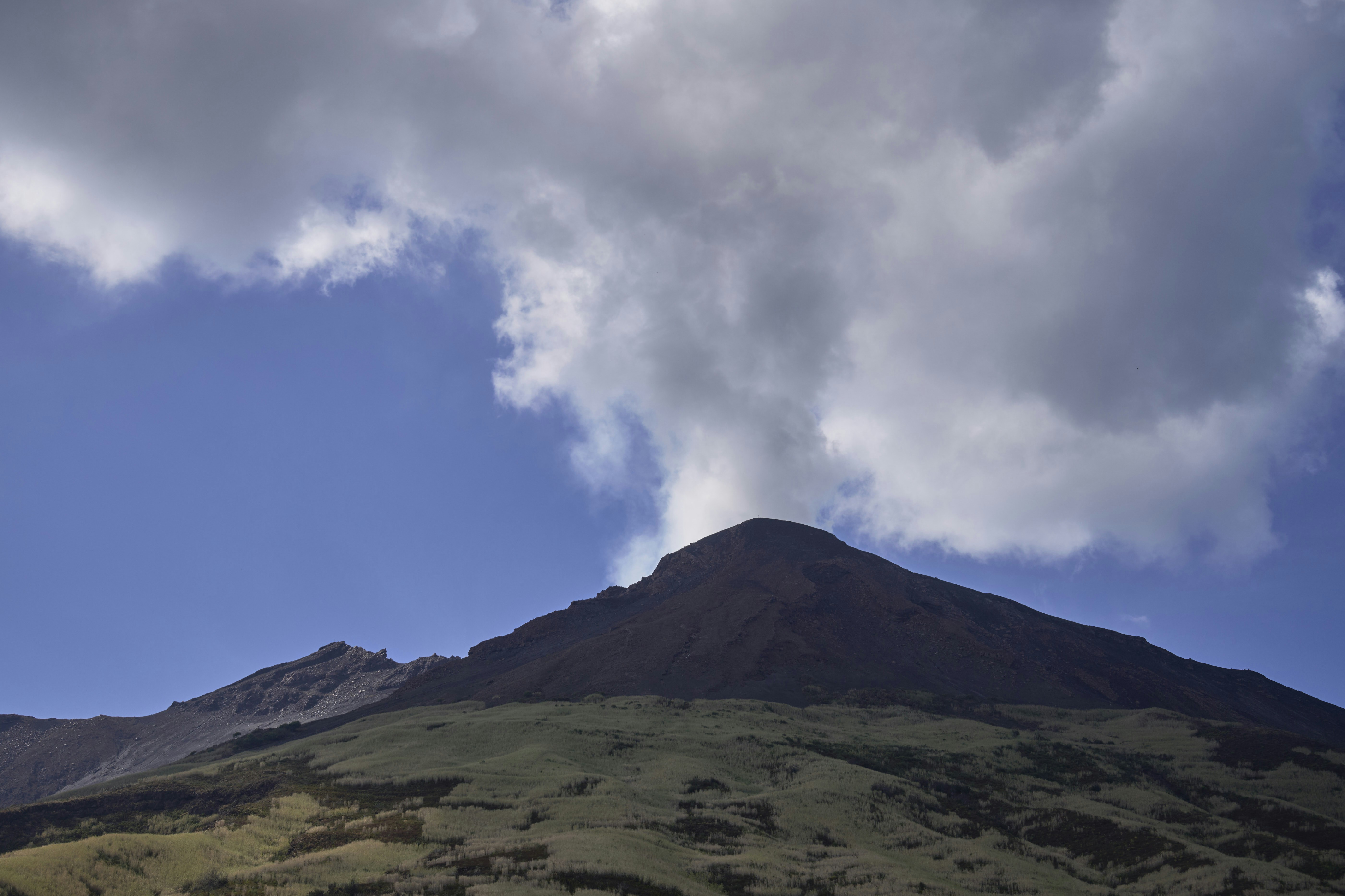 Volcano emitting a plume of smoke against a clear blue sky, with verdant slopes contrasting the rugged peak.