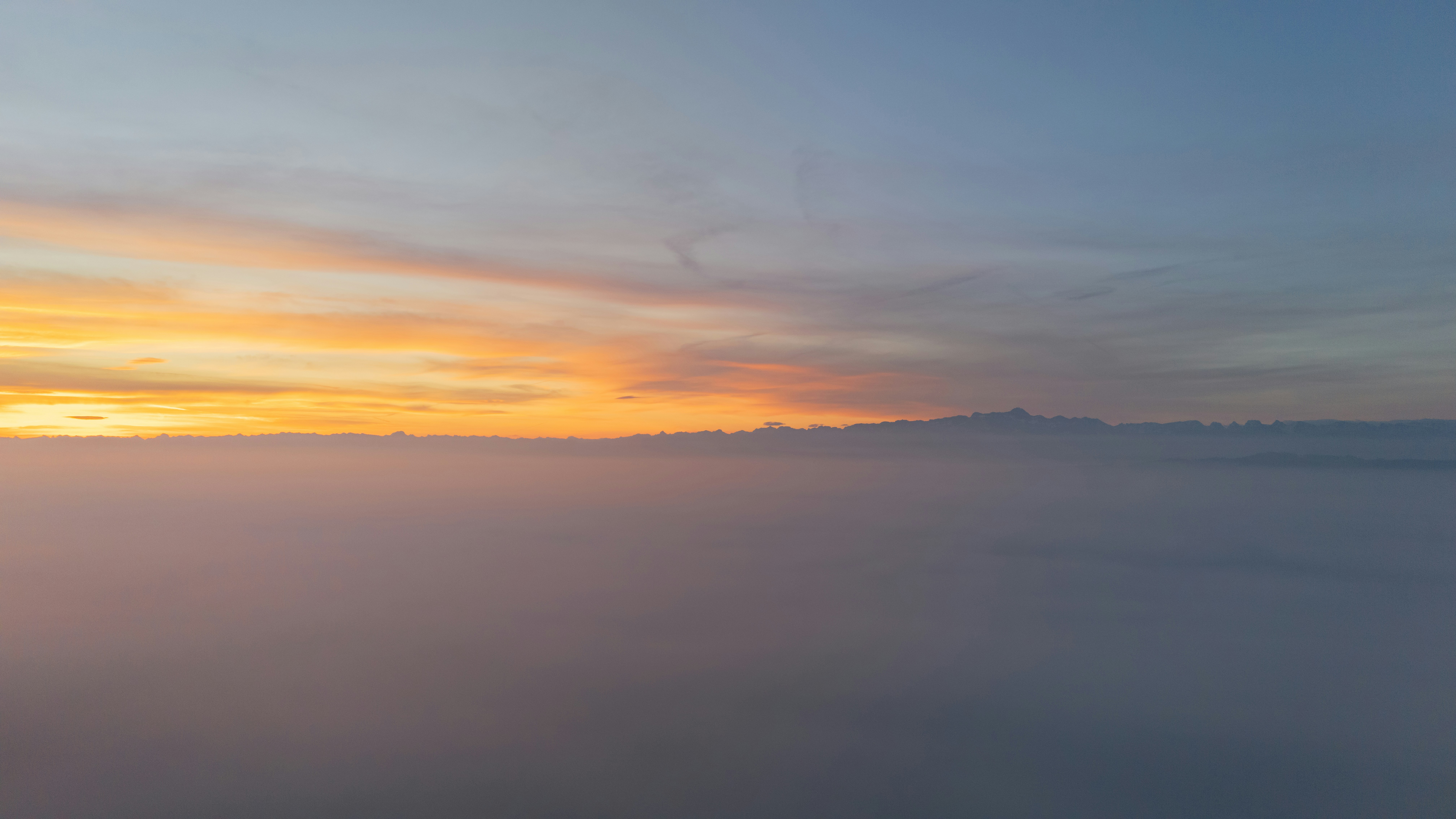 drone view of a breathtaking sunrise above a sea of fog, with the distant silhouette of mountain peaks along the horizon. The sky glows in soft pastel hues of orange, yellow, and purple, gently transitioning into the cool blue tones of early morning. The tranquil atmosphere and the delicate gradient of colors create a serene and dreamlike scene that captures the beauty of nature at dawn. | Sunrise over a misty mountain range.