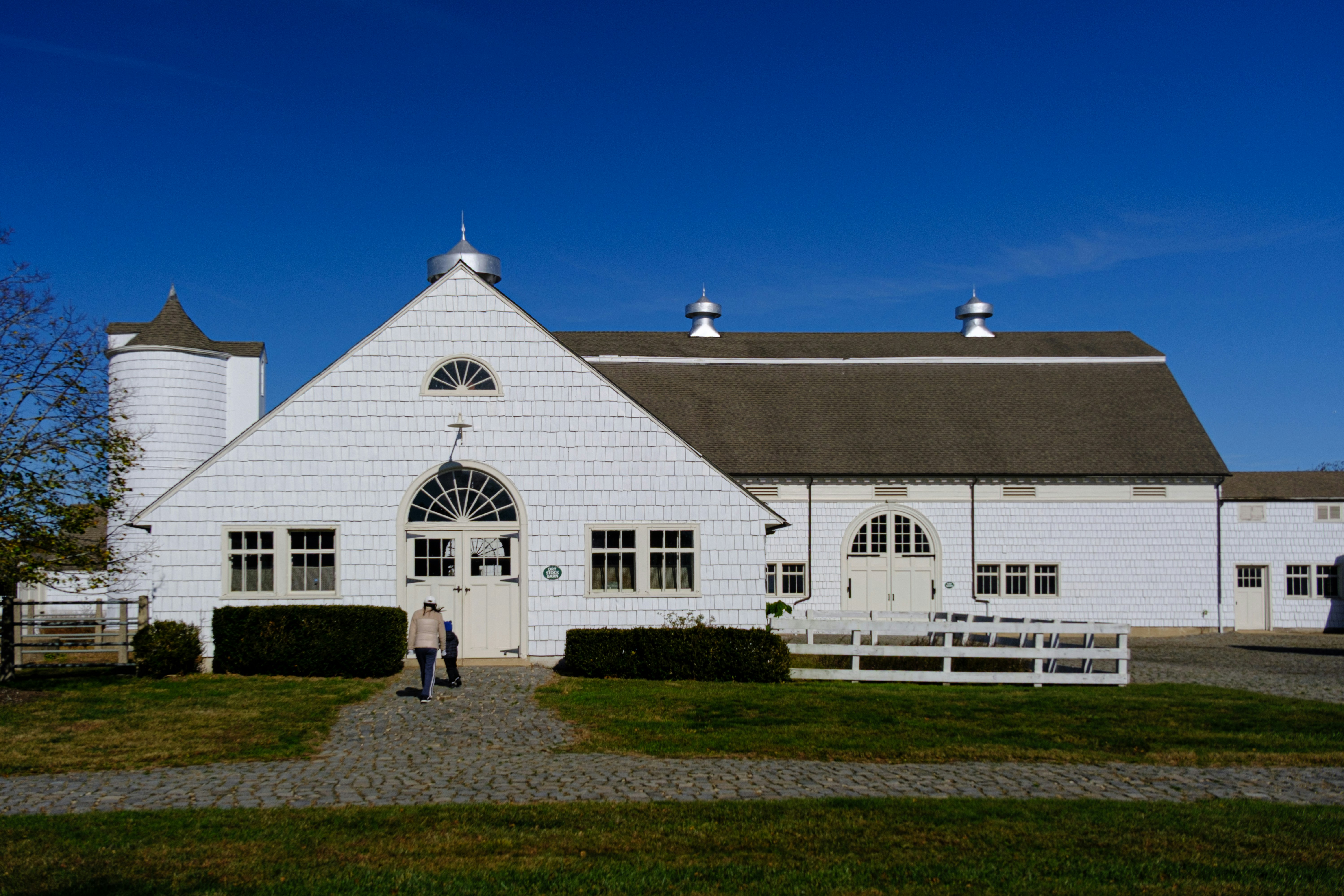 A historic barn with white shingle siding and a silo, set against a clear blue sky, showcasing the serene countryside. A person walks towards the entrance.