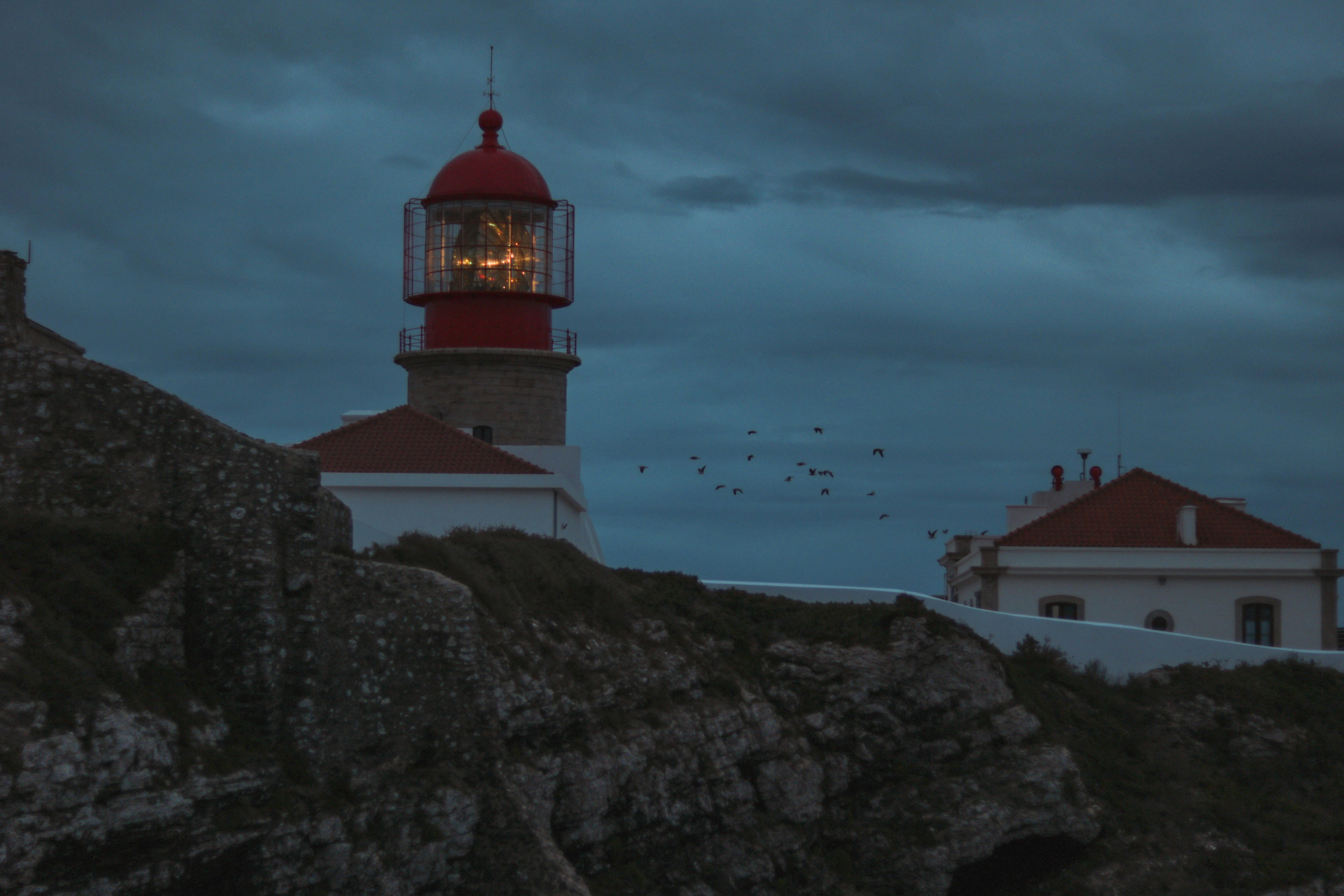Lighthouse standing resilient against a darkening sky, with birds flying nearby and a warm light glowing from its lantern.