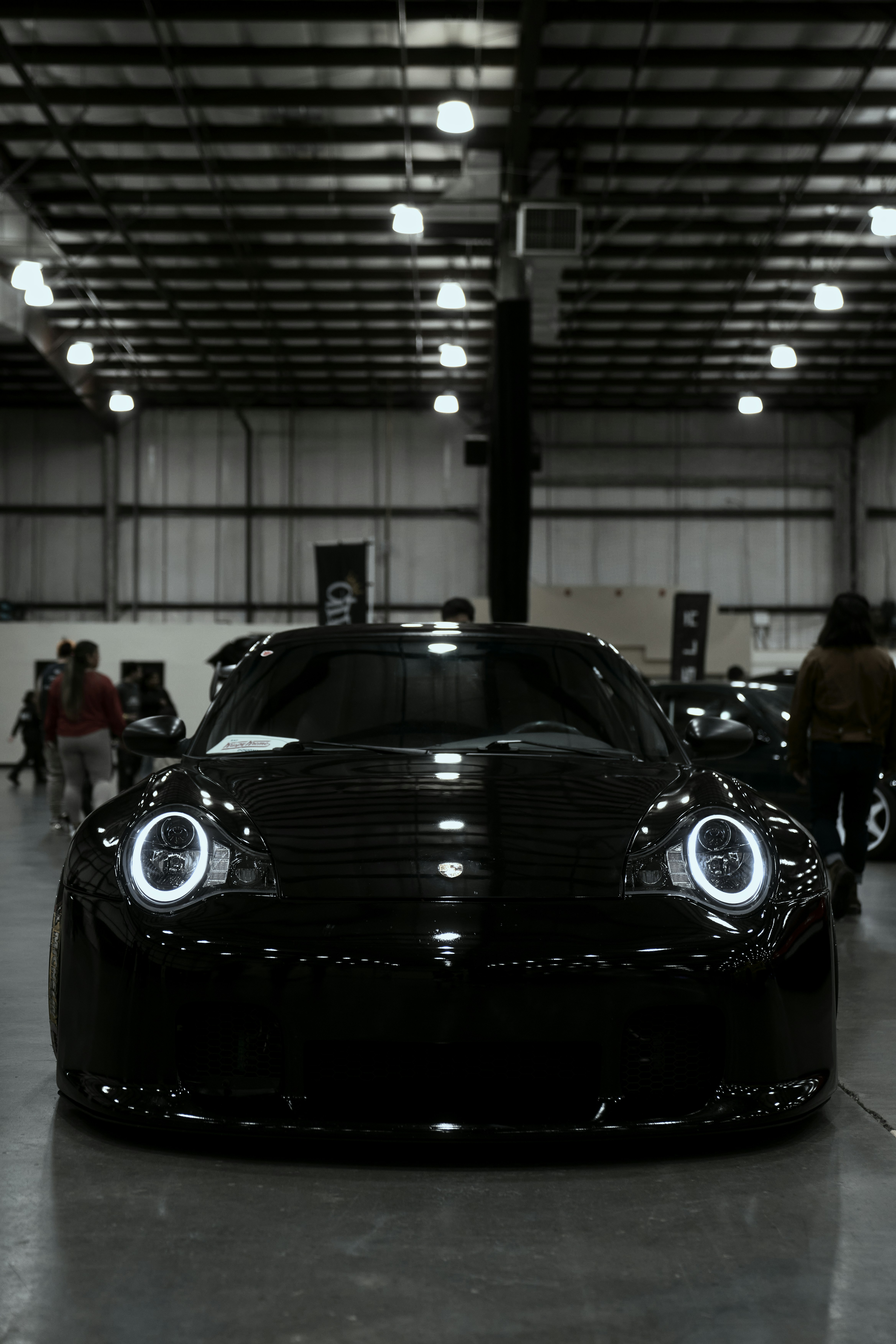 Sleek black sports car with striking headlights showcased in a dimly lit exhibition hall, surrounded by onlookers.