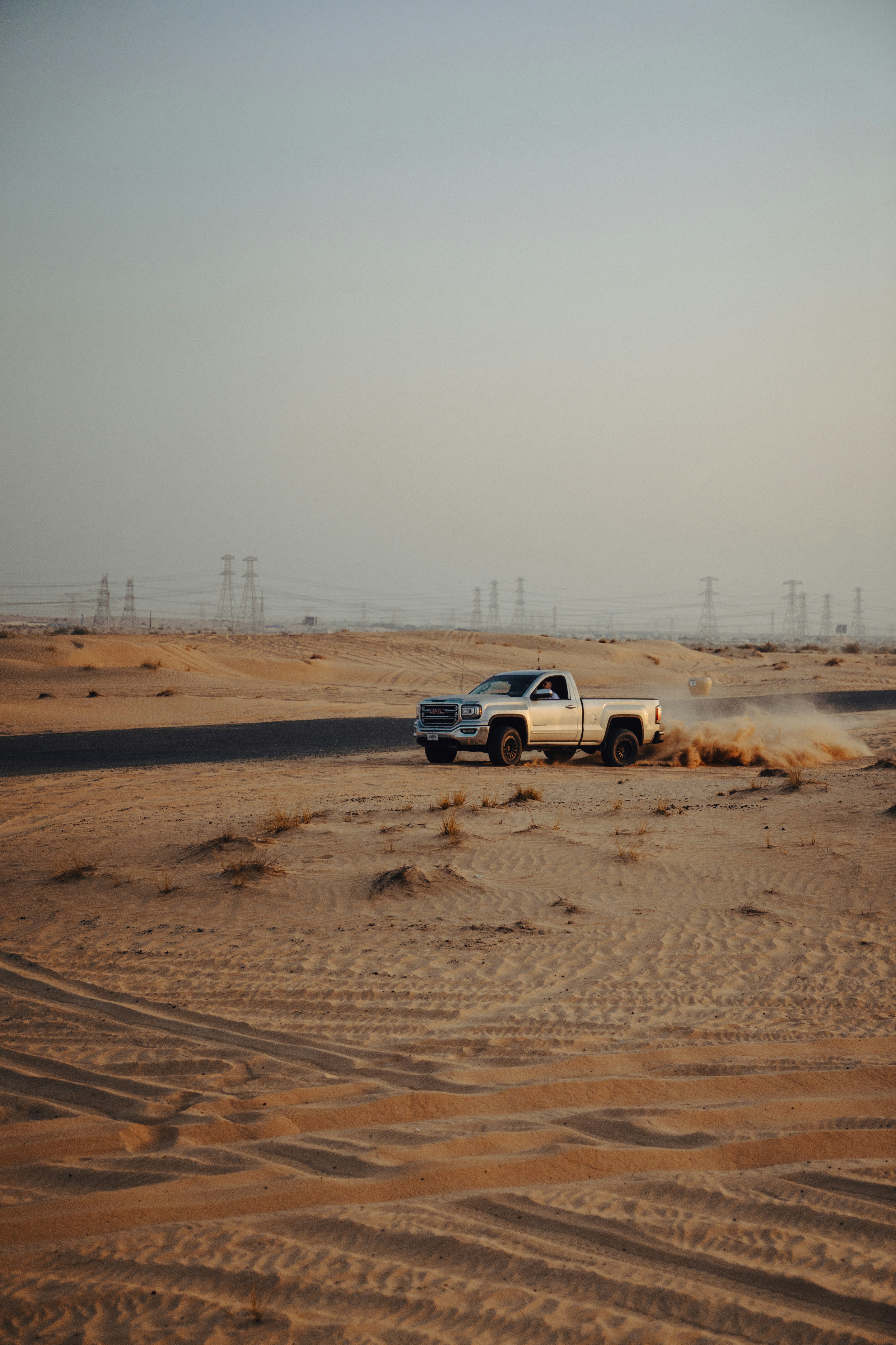 A white pickup truck kicks up sand while navigating a winding road through a vast desert landscape, framed by distant power lines.