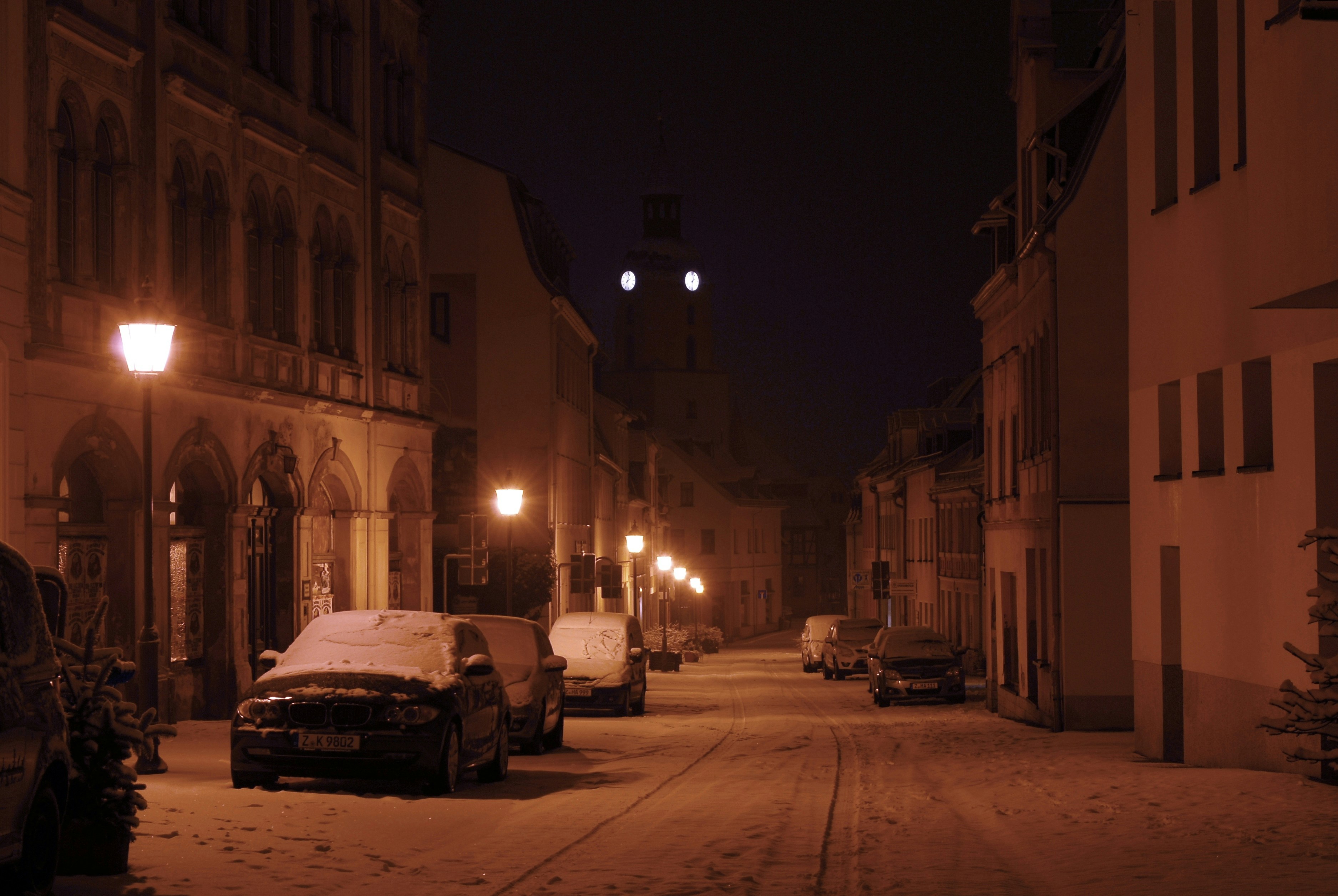 Snow-covered street at night, illuminated by warm street lamps, with parked cars lining the road and a clock tower in the background.