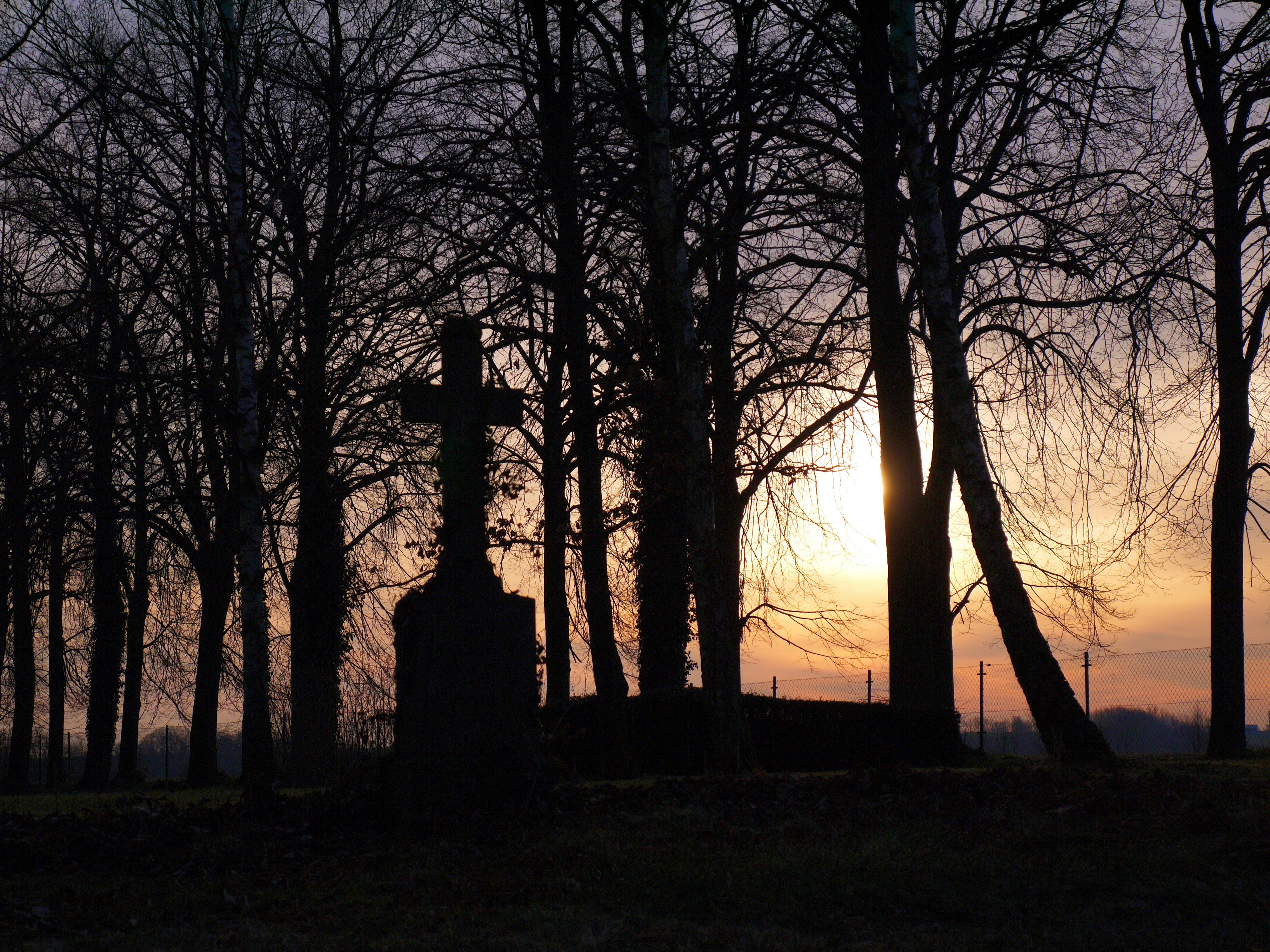 Silhouetted cross stands amidst bare trees against a glowing sunset backdrop, evoking a sense of reflection and tranquility.