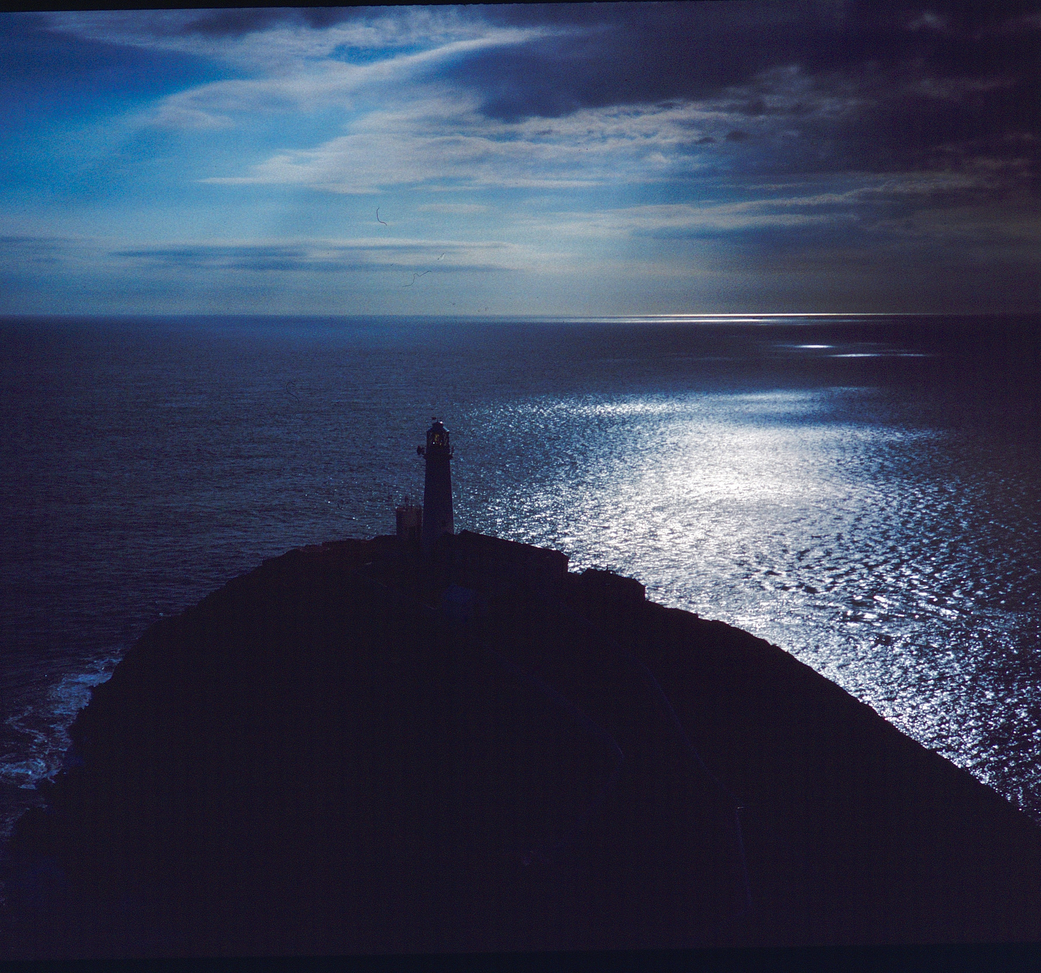 Lighthouse perched on a rocky outcrop, casting its light over tranquil ocean waters reflecting the sky's hues.