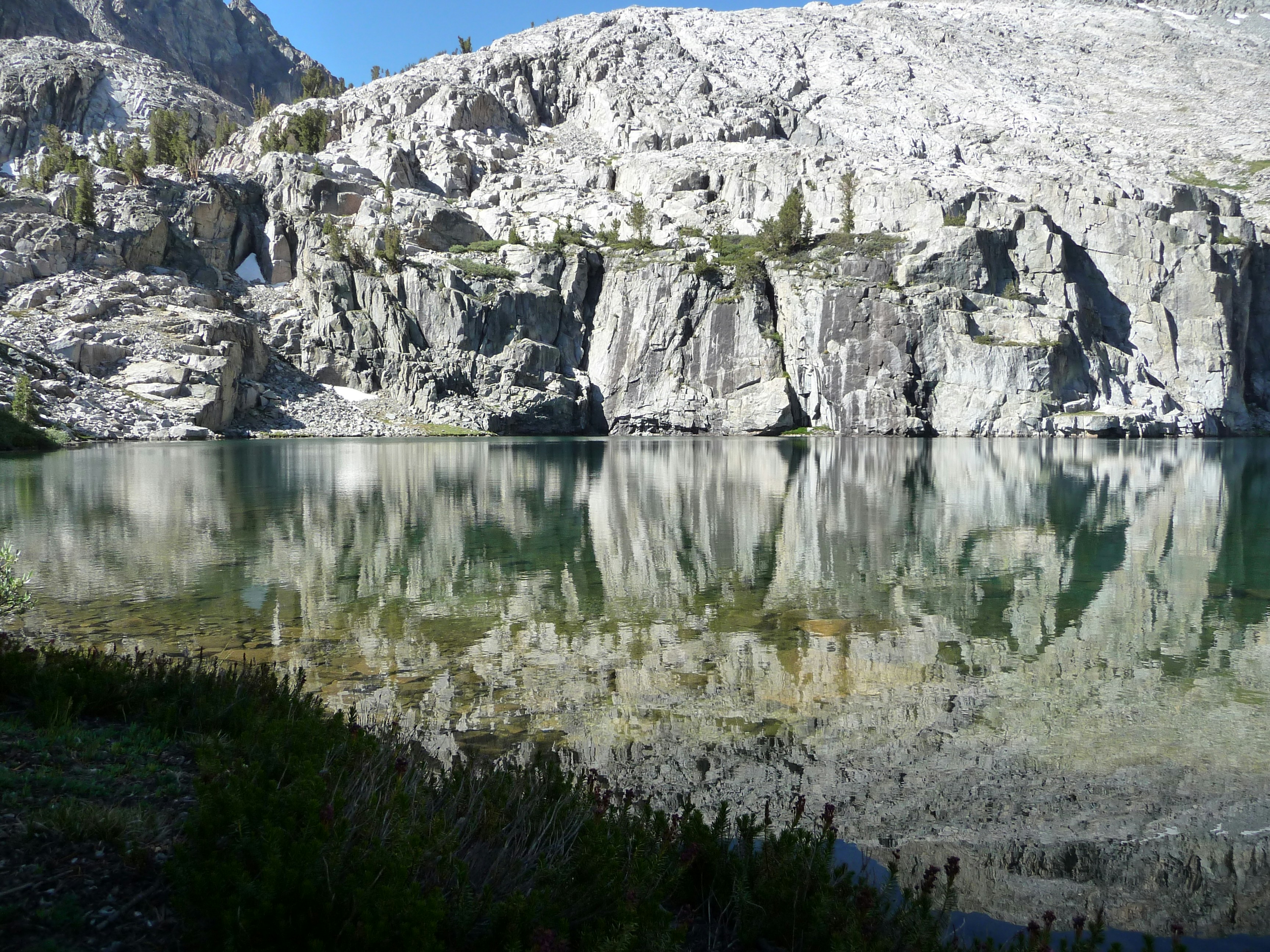 Crystal-clear mountain lake reflecting rugged cliffs and blue sky, surrounded by lush greenery.