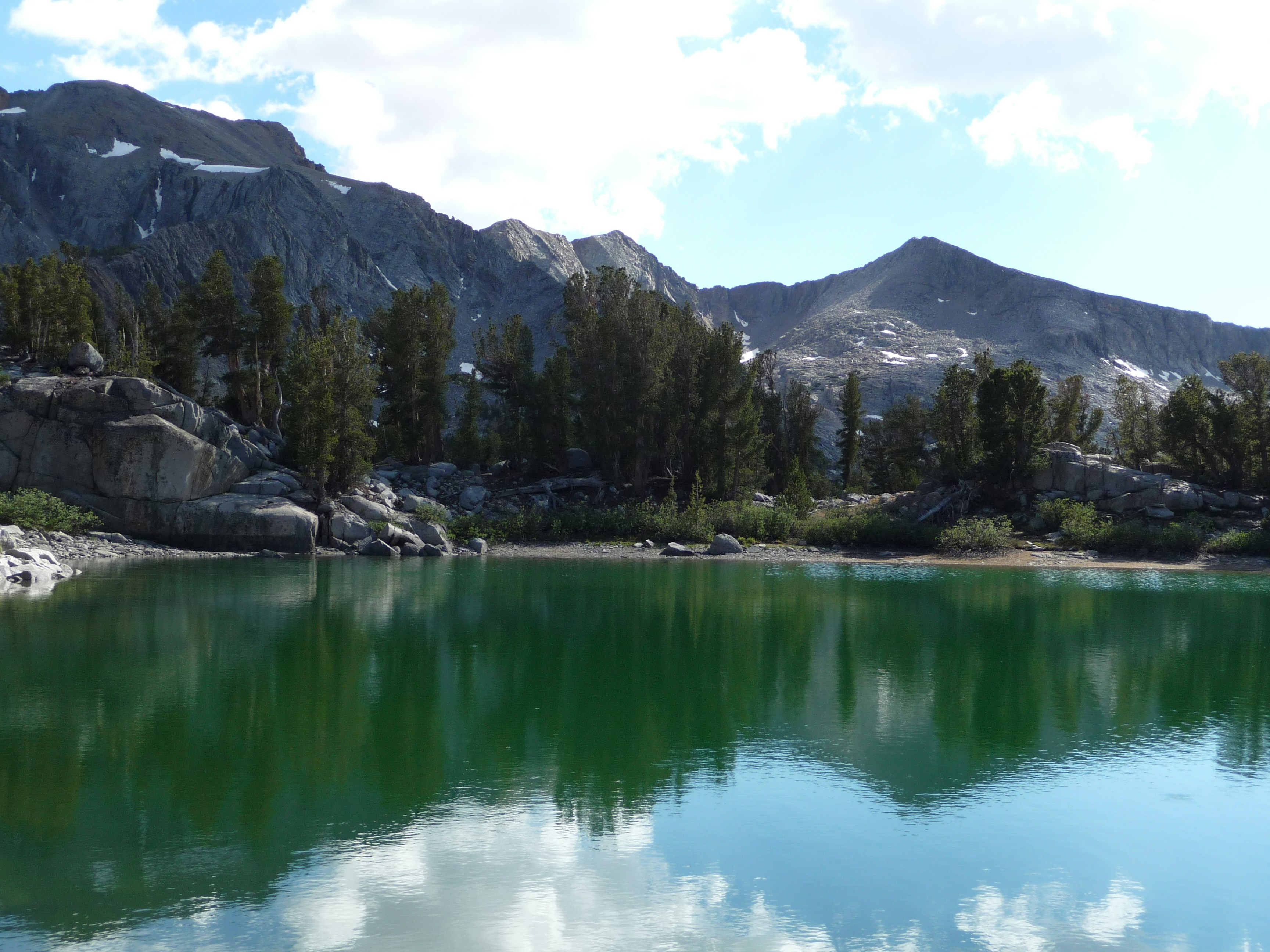 Tranquil alpine lake reflecting rocky mountains and lush trees under a bright sky.