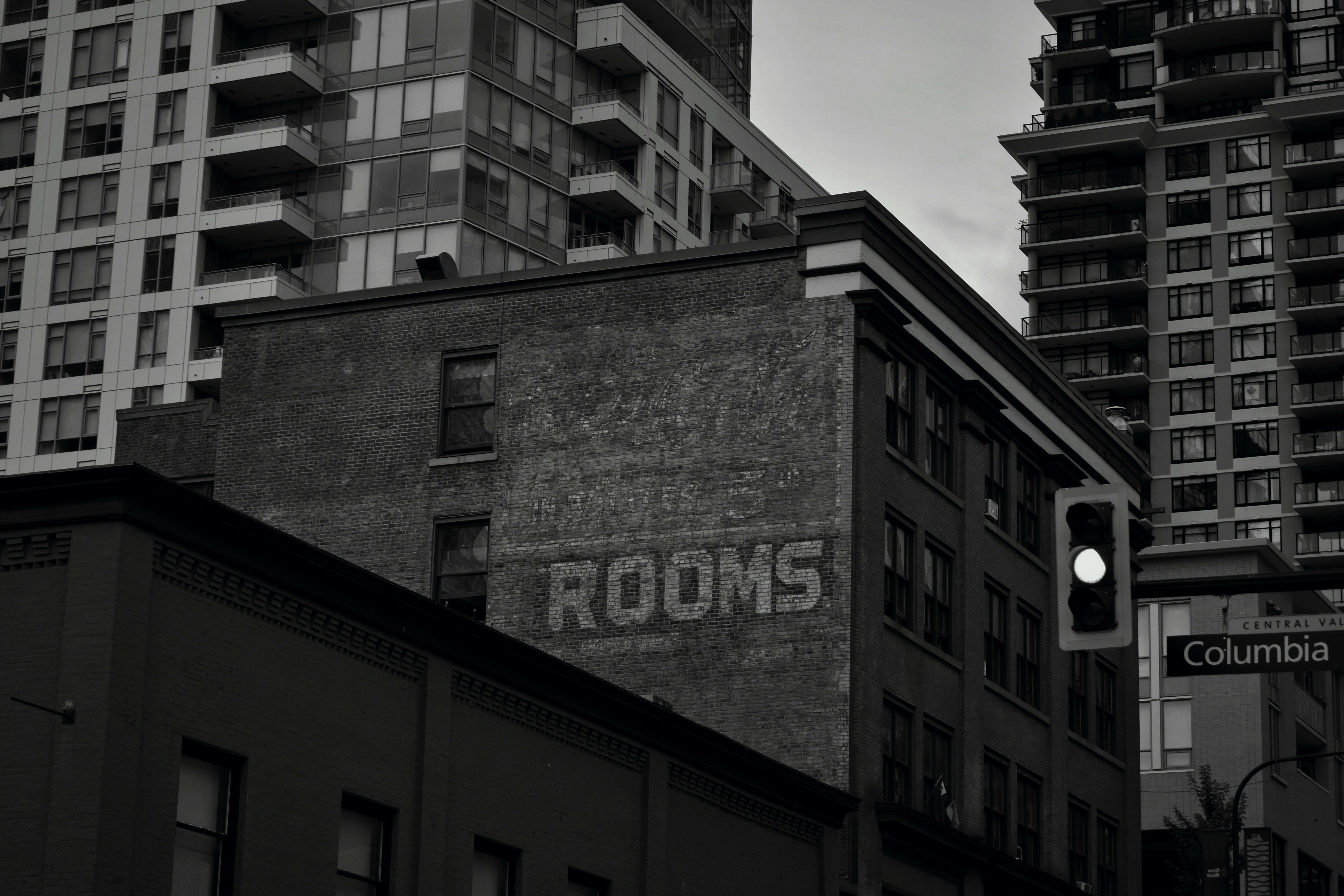 Faded advertisement for 'Rooms' on a historic building juxtaposed against modern high-rises. The monochrome palette enhances the contrast between old and new.