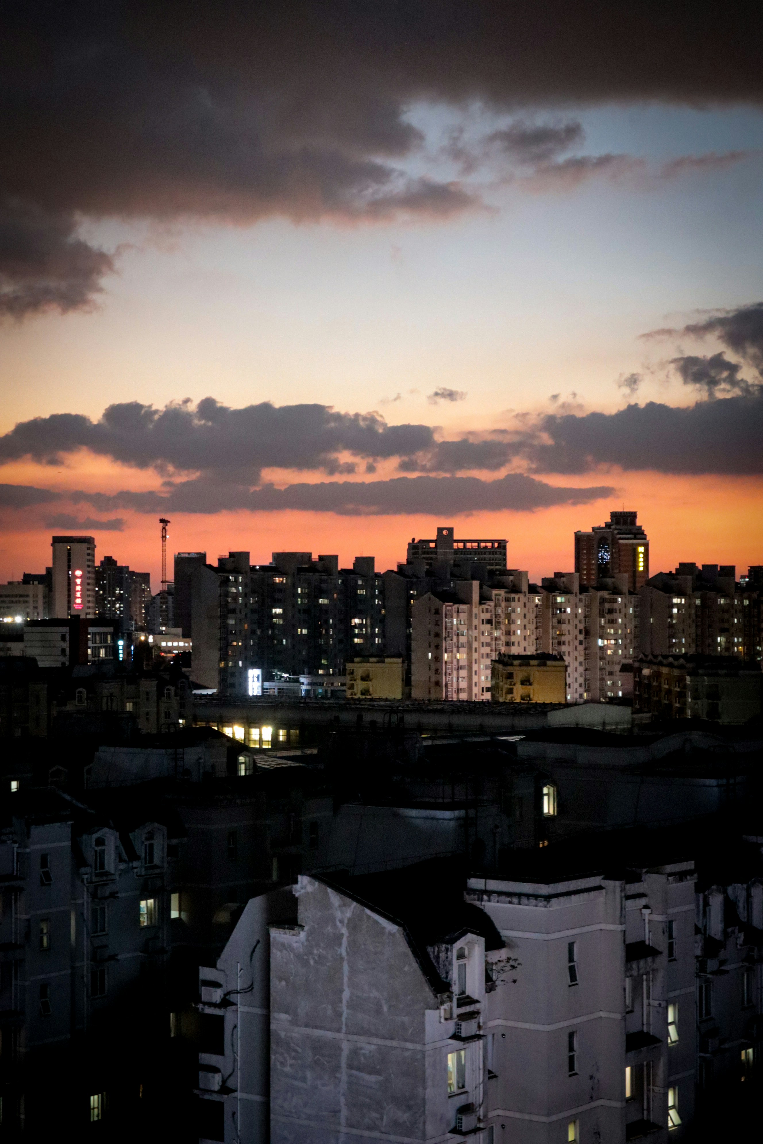 City skyline illuminated by the warm glow of sunset, contrasting with darkened rooftops below. The scene captures the transition from day to night.