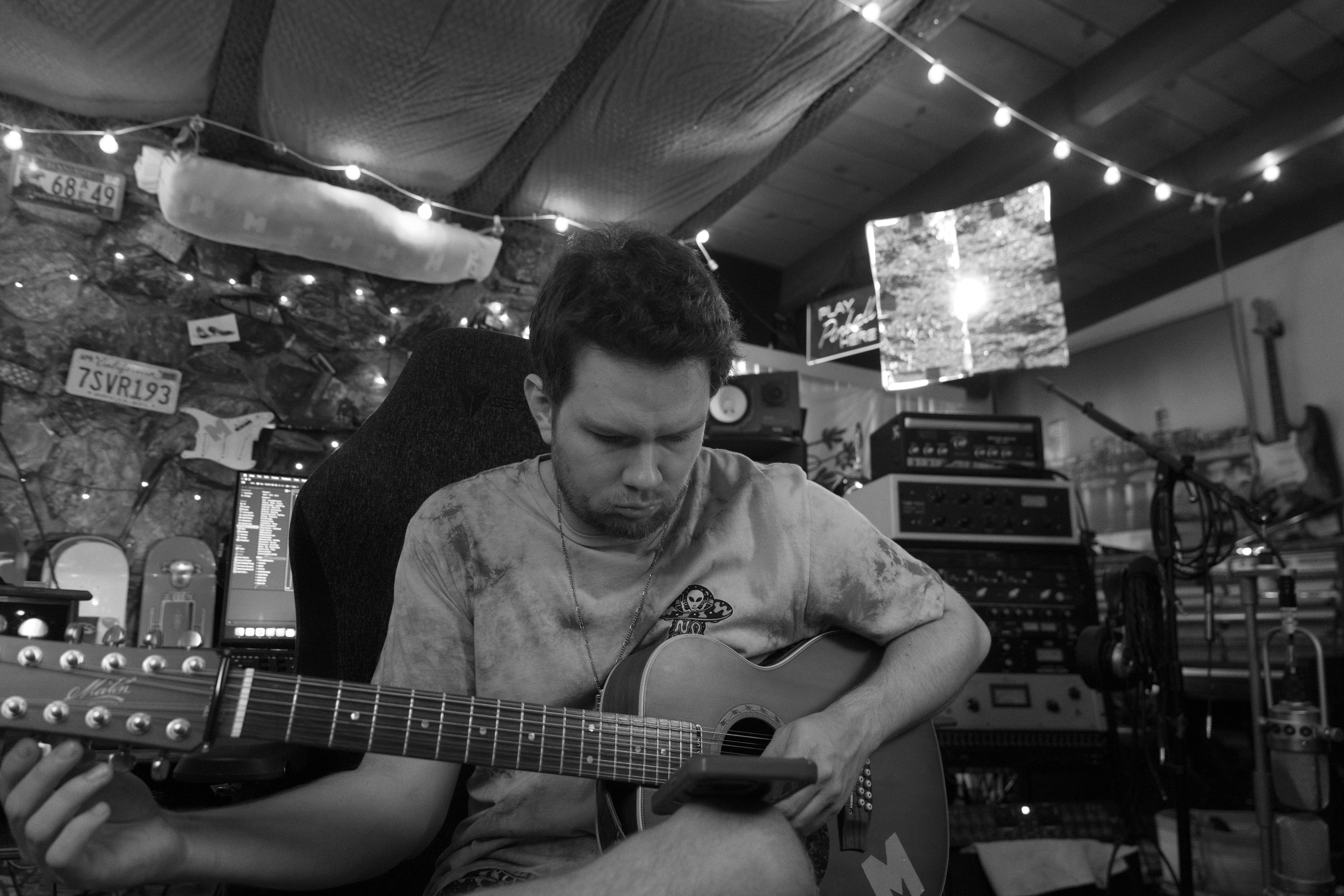 Musician focused on a smartphone while playing an acoustic guitar in a cozy, well-equipped studio filled with lights and musical gear.