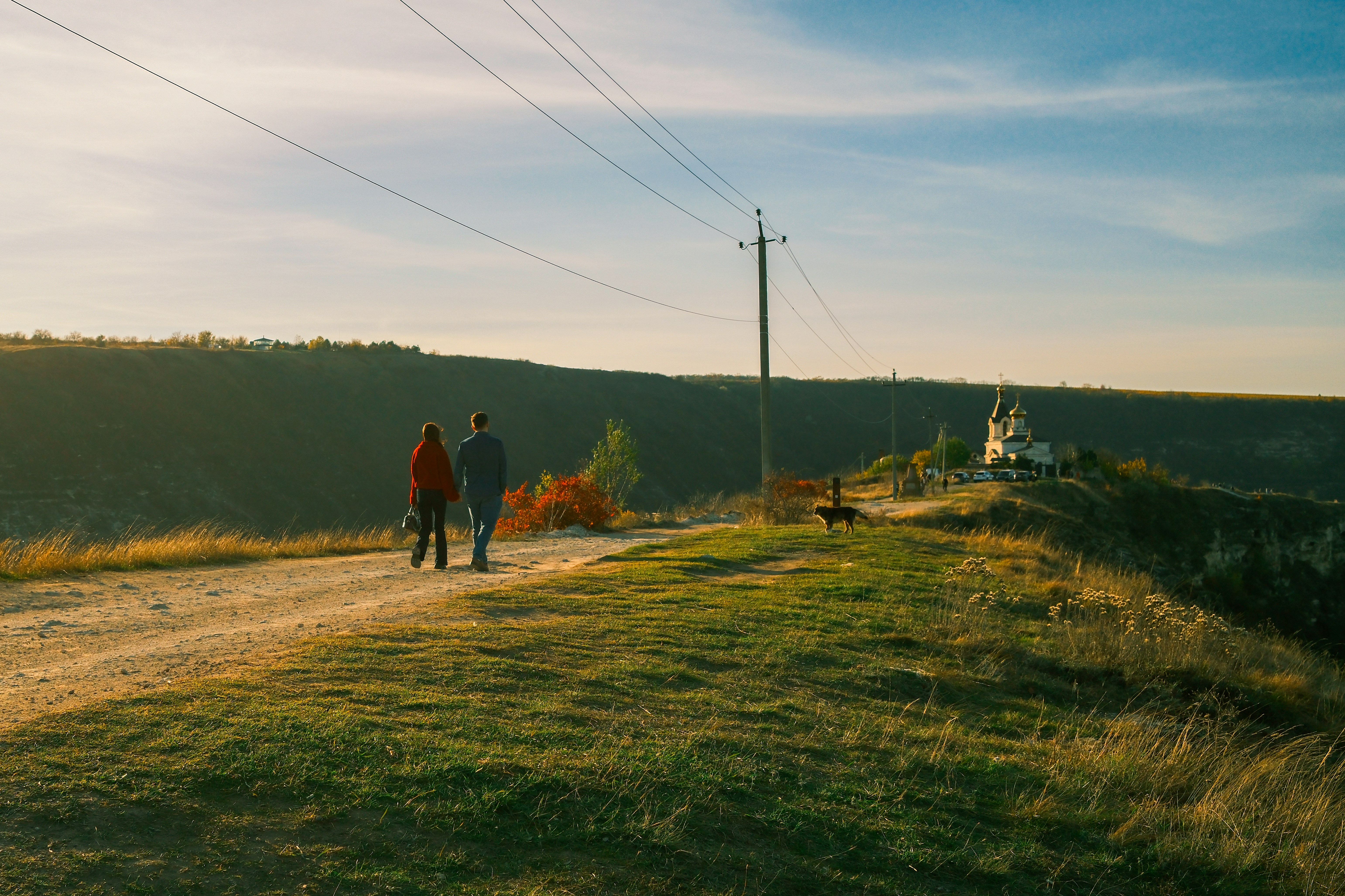 Couple walking hand-in-hand along a dirt path atop a cliff, with a distant building and dog in view. The scene captures the tranquility of an evening walk amidst nature.