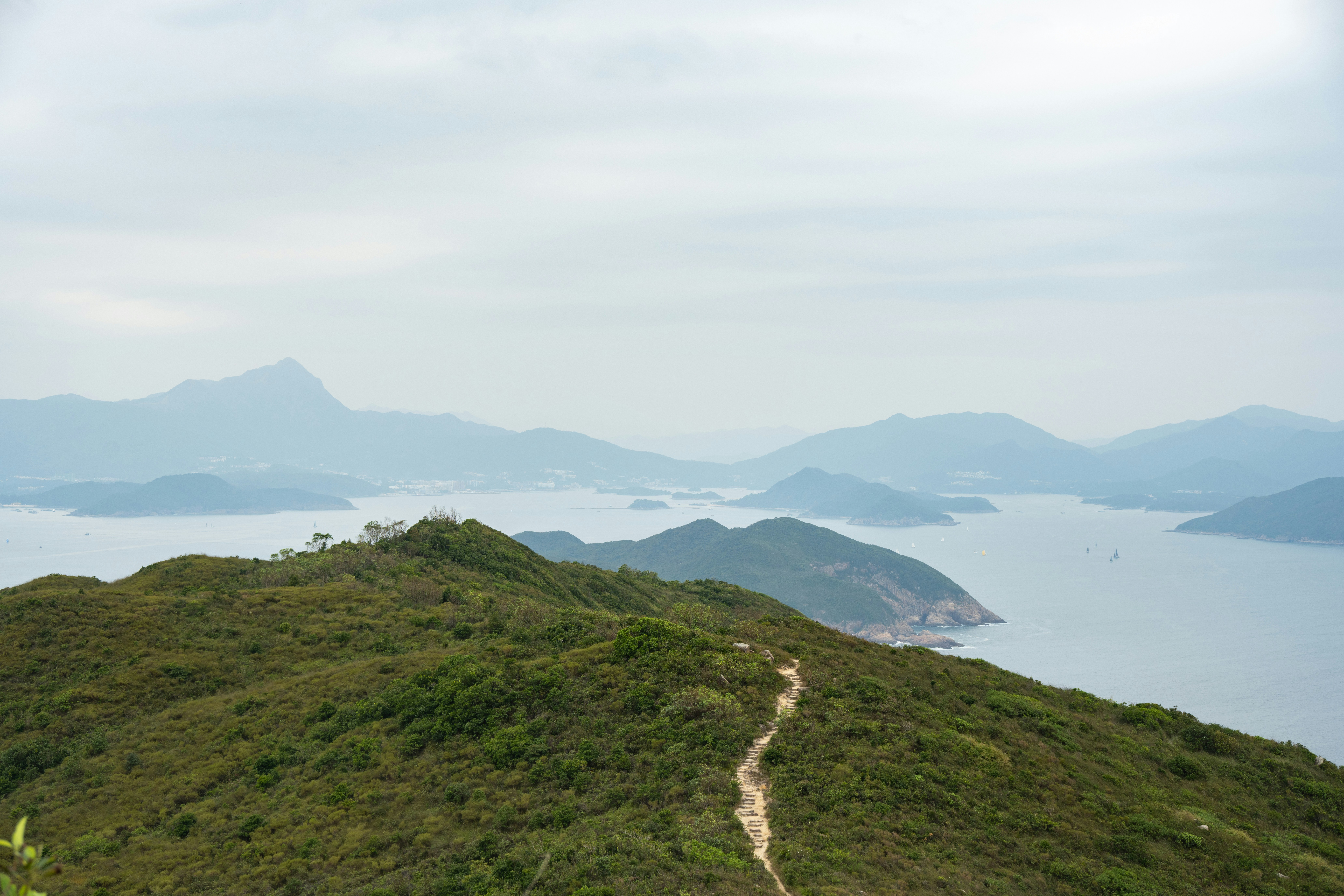Lush green hills lead to a tranquil bay dotted with islands under a soft, overcast sky. The winding path invites exploration.