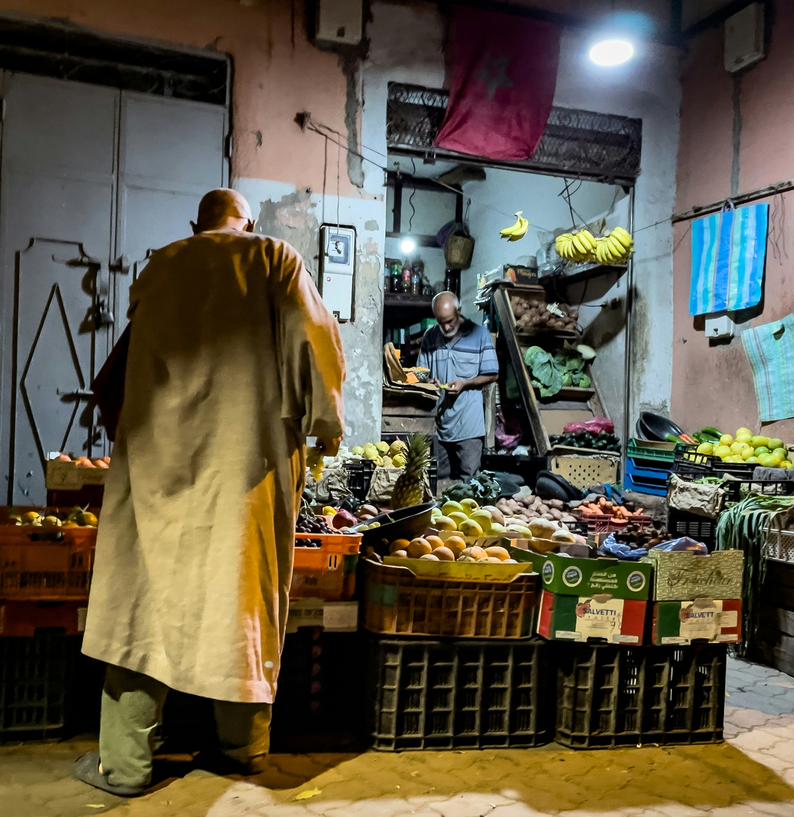 A bustling fruit and vegetable market at night, illuminated by soft overhead lights, showcasing vibrant produce and shoppers engaged in lively transactions.