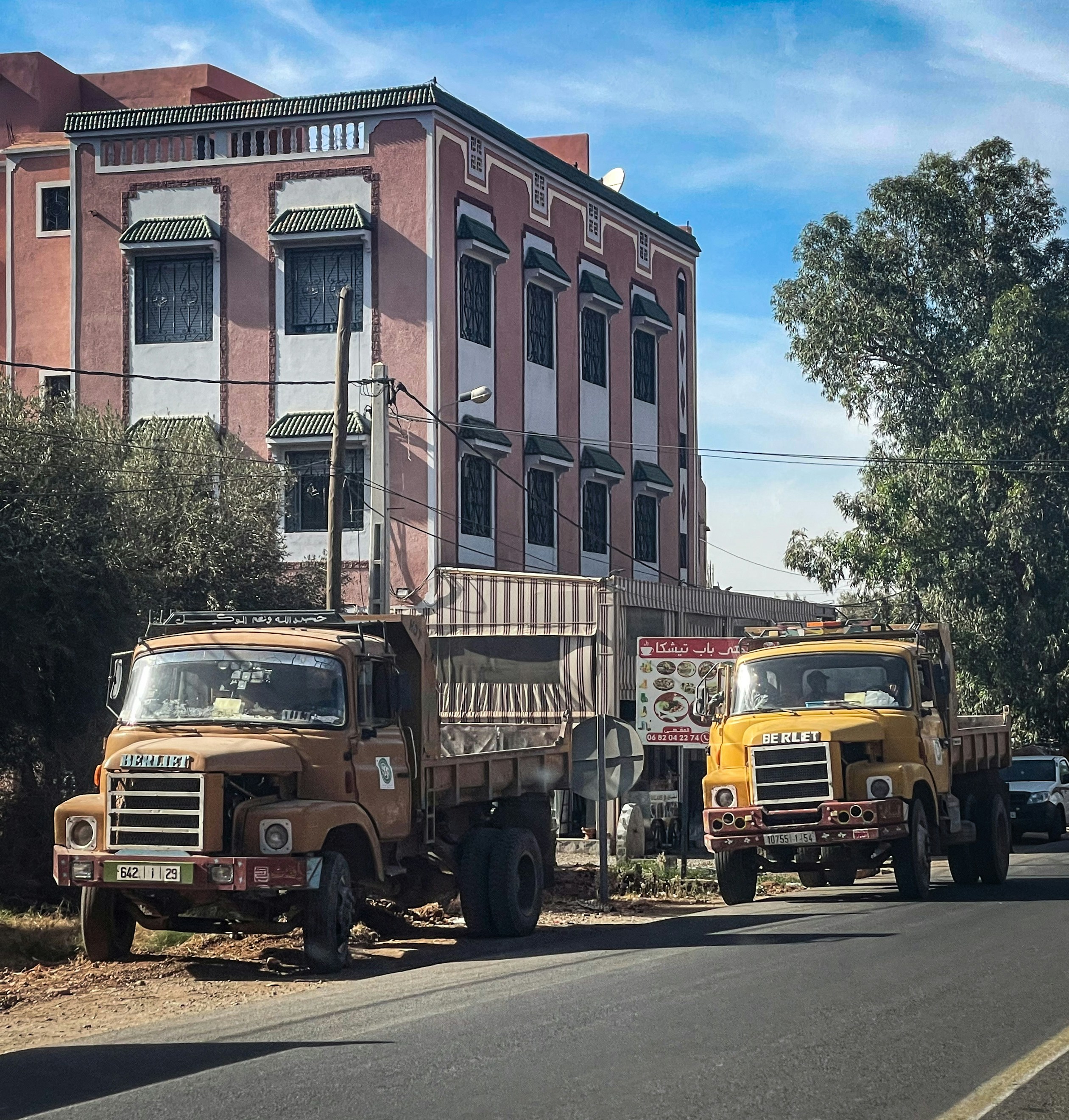 Two vintage trucks parked alongside a road, framed by a colorful building and greenery, capturing the essence of everyday life in an urban setting.