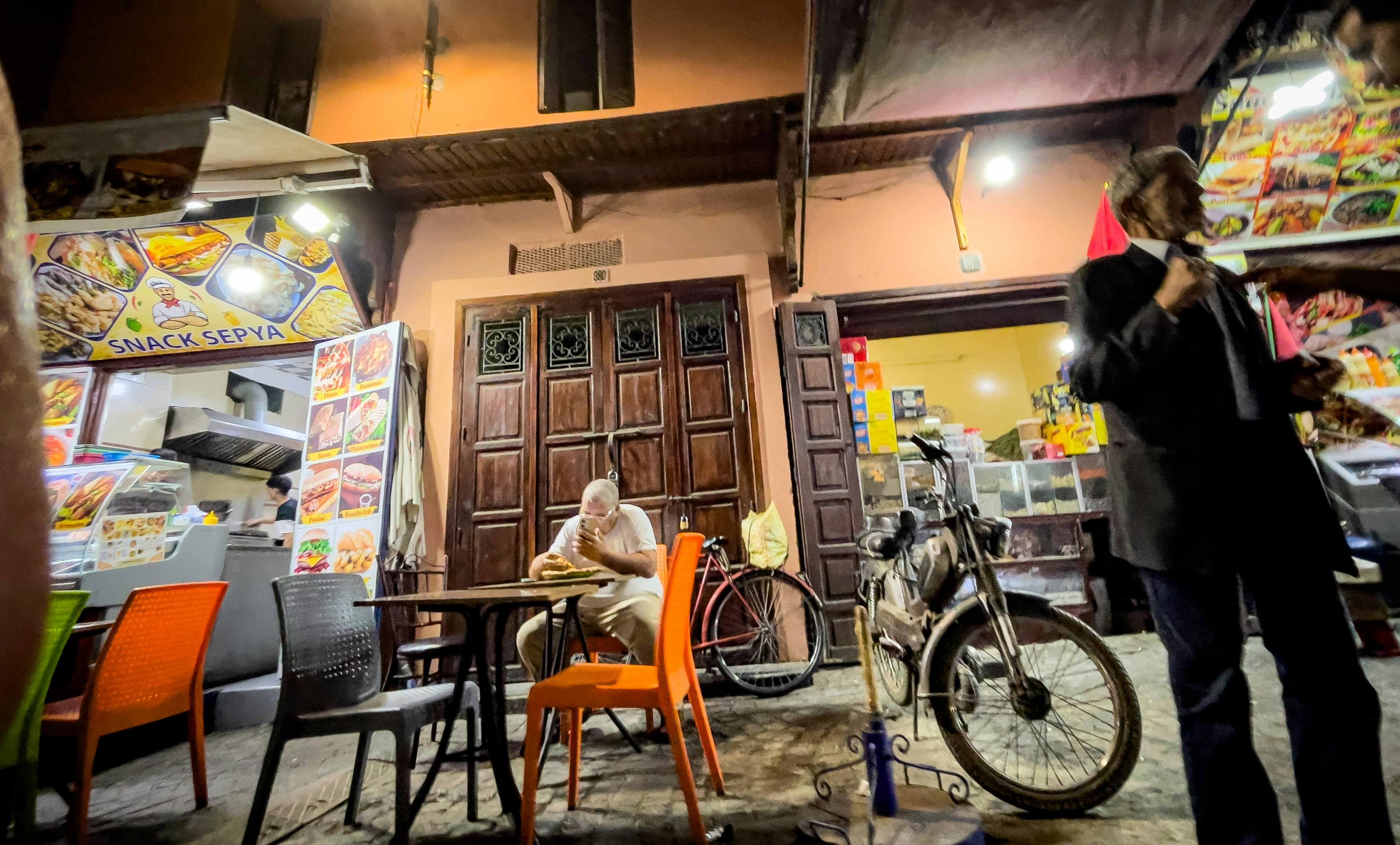 A relaxed diner enjoys a meal at a bustling food stall while a cyclist waits nearby. The colorful ambiance highlights the lively street atmosphere.