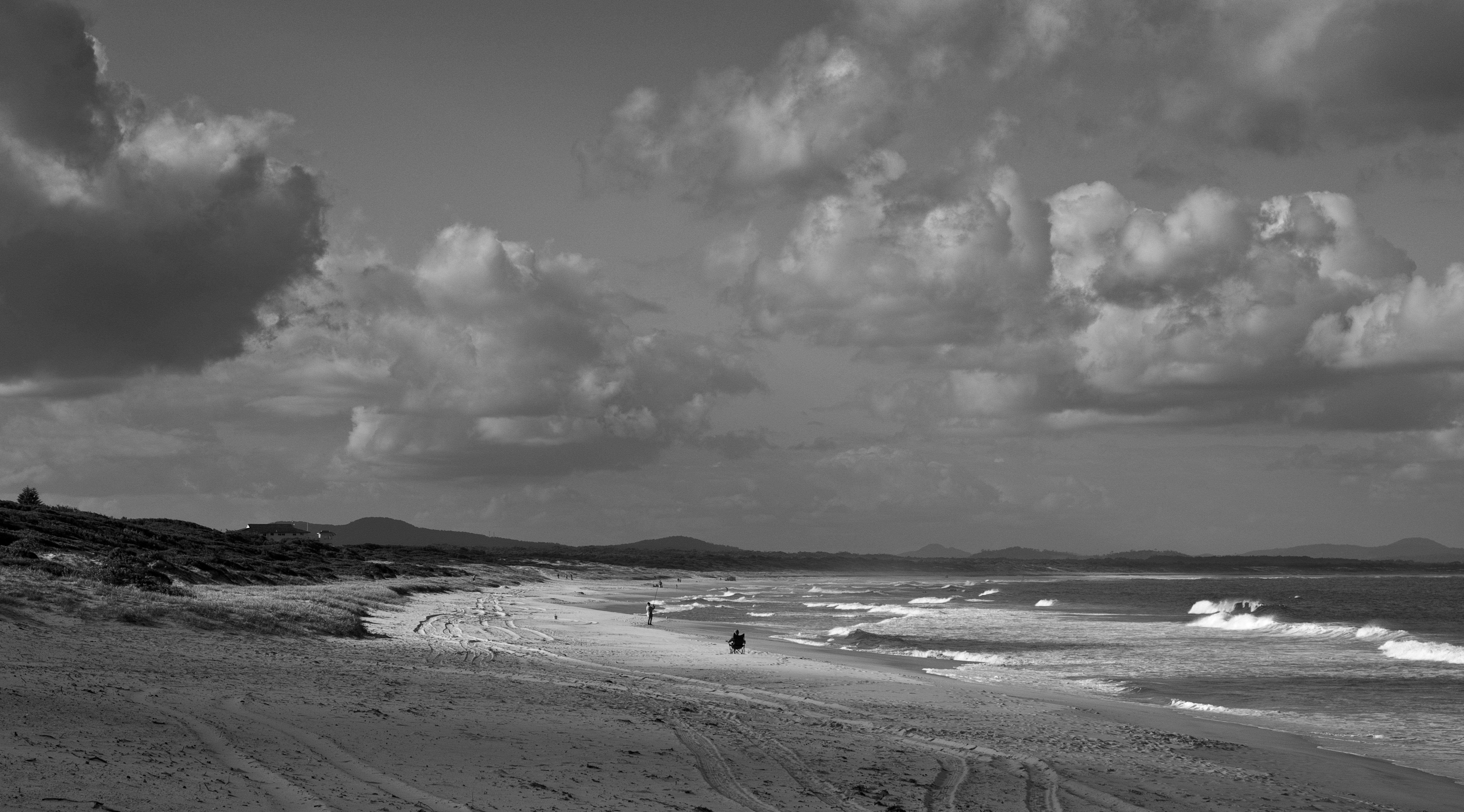 Lonely beach scene under a dramatic sky with gentle waves and distant figures walking along the shore.