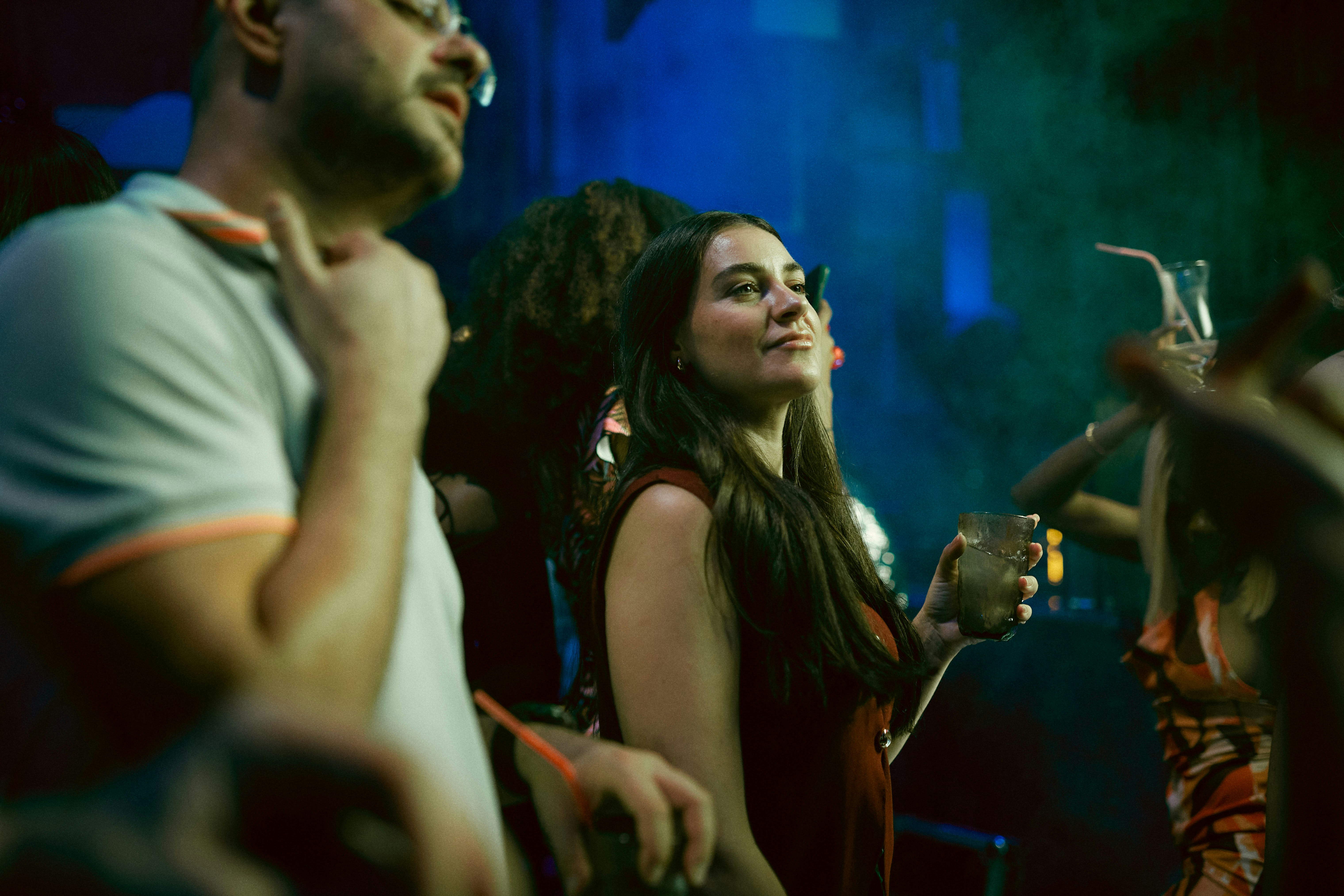 A woman enjoying a drink in a lively nightlife setting, surrounded by friends and vibrant lights. The atmosphere is electric with energy.