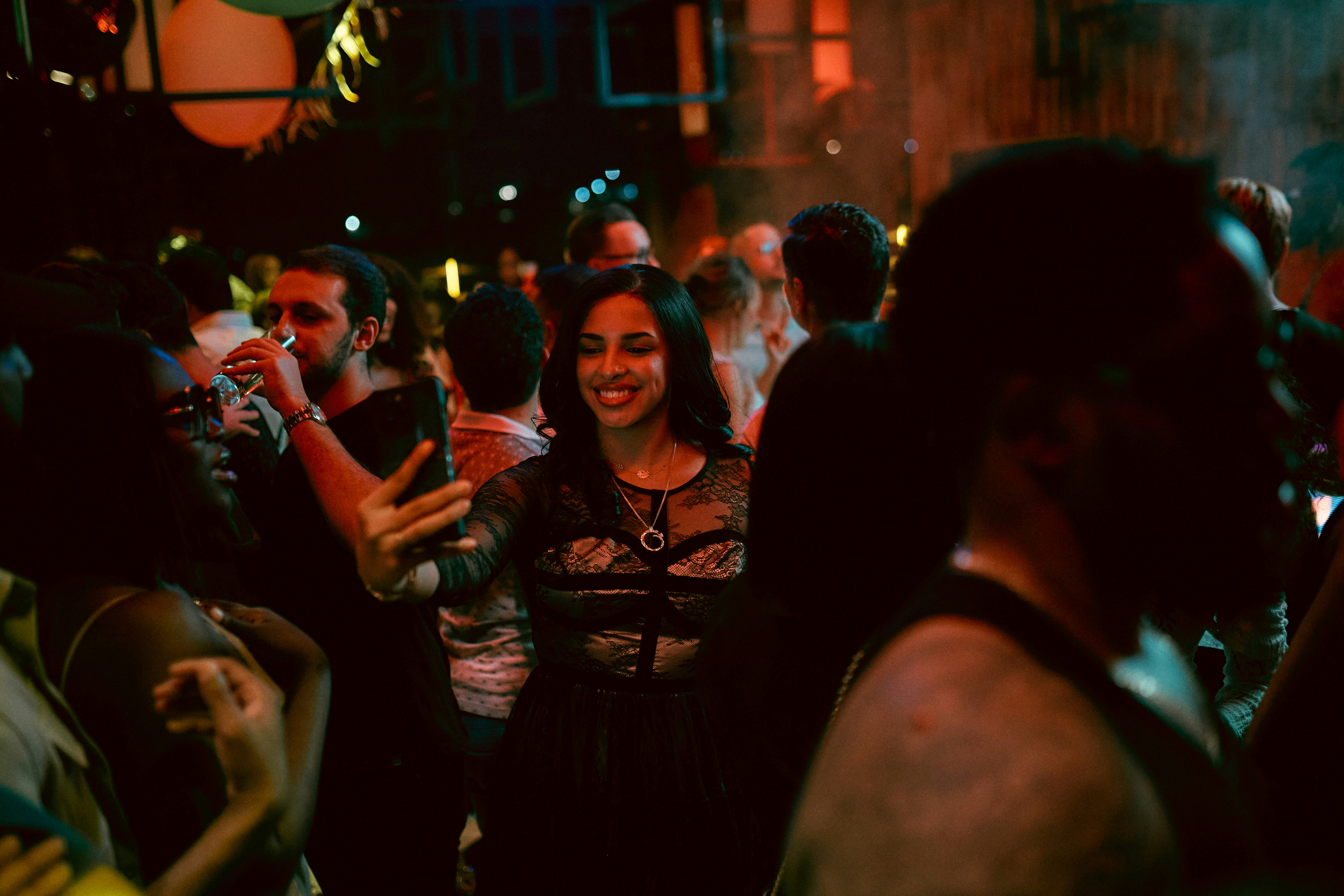 A lively gathering in a vibrant atmosphere, featuring people dancing and enjoying the night. The focus is on a woman in a black dress, exuding joy amidst the crowd.