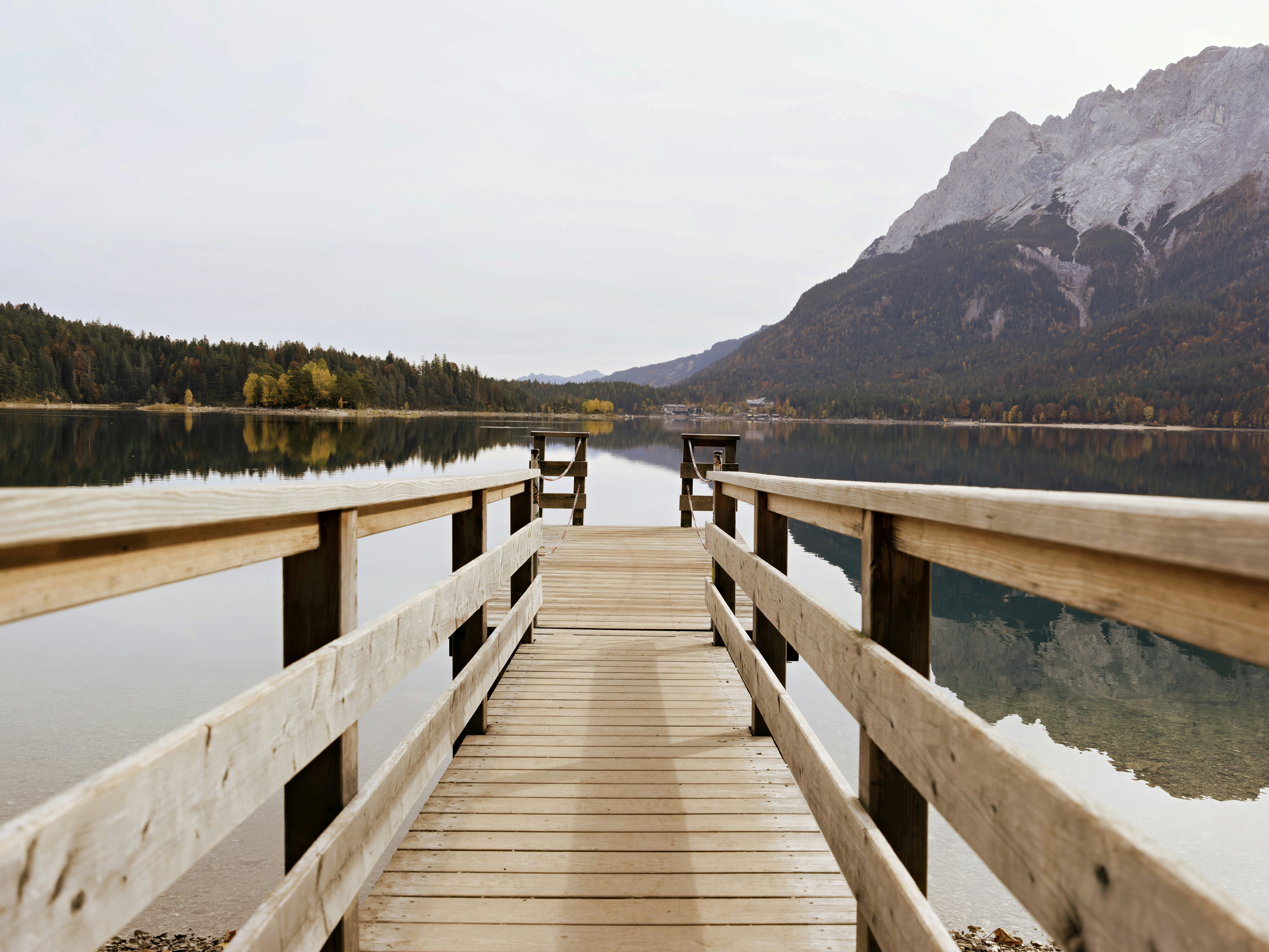 Wooden dock extending into a serene lake, framed by mountains and autumn foliage. Still waters reflect the surrounding landscape.