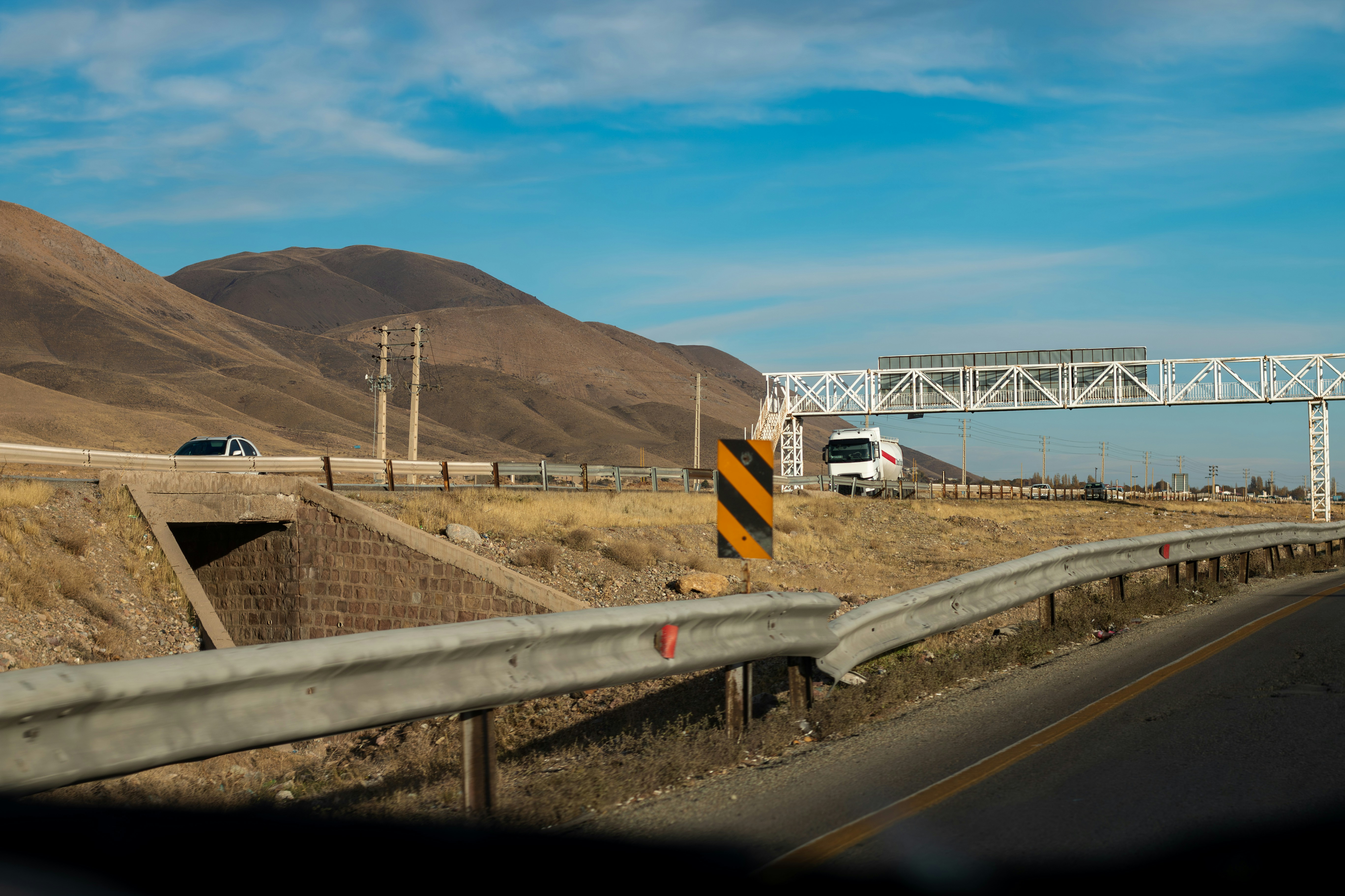 Desert highway winding through rolling hills under a clear blue sky, featuring a bridge overhead and vehicles on the road.