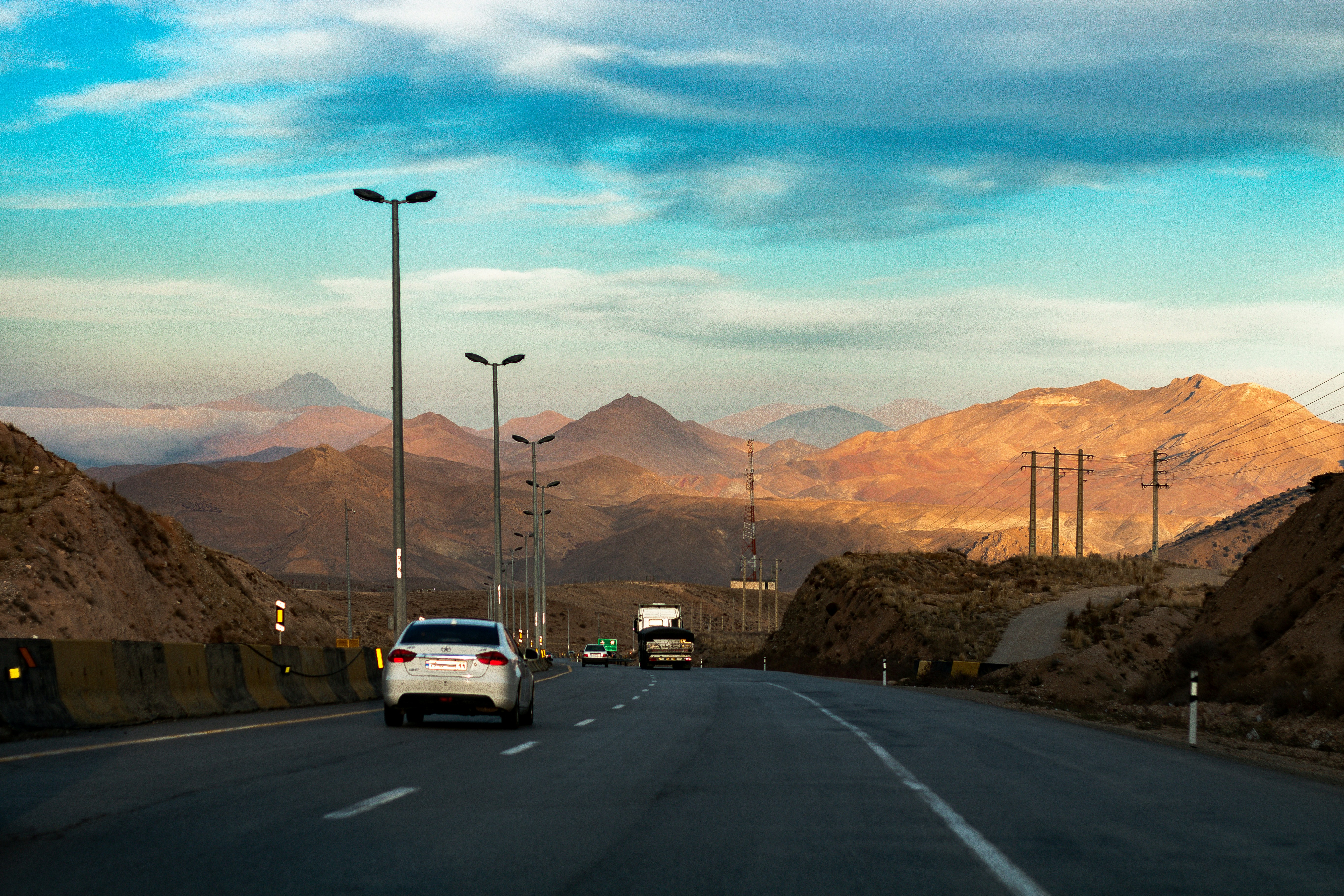 A winding road leads through rugged mountains, with vehicles traversing the landscape under a softly lit sky.