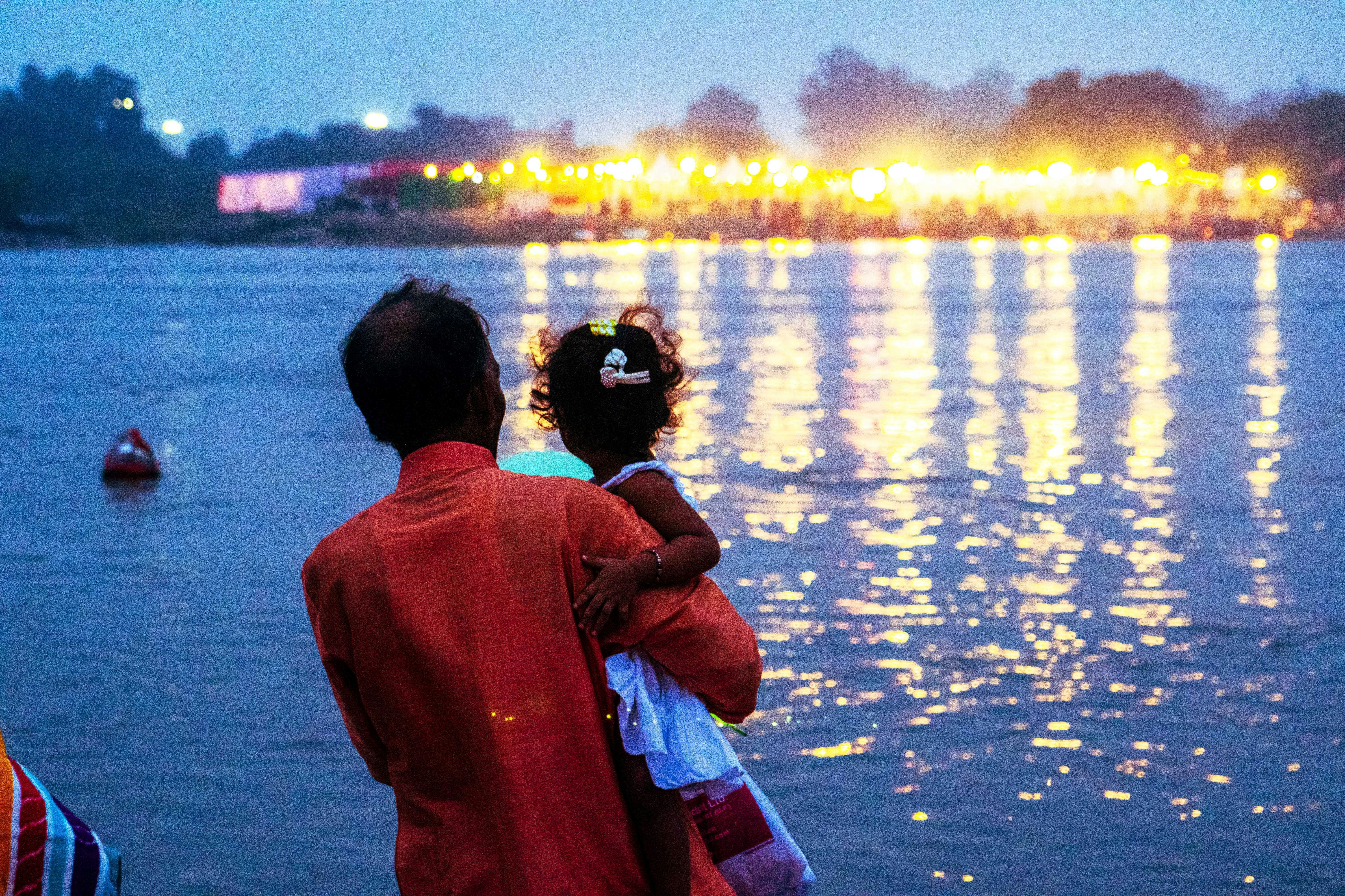 A father holds his child as they gaze at the shimmering lights reflecting on the water during an evening celebration.