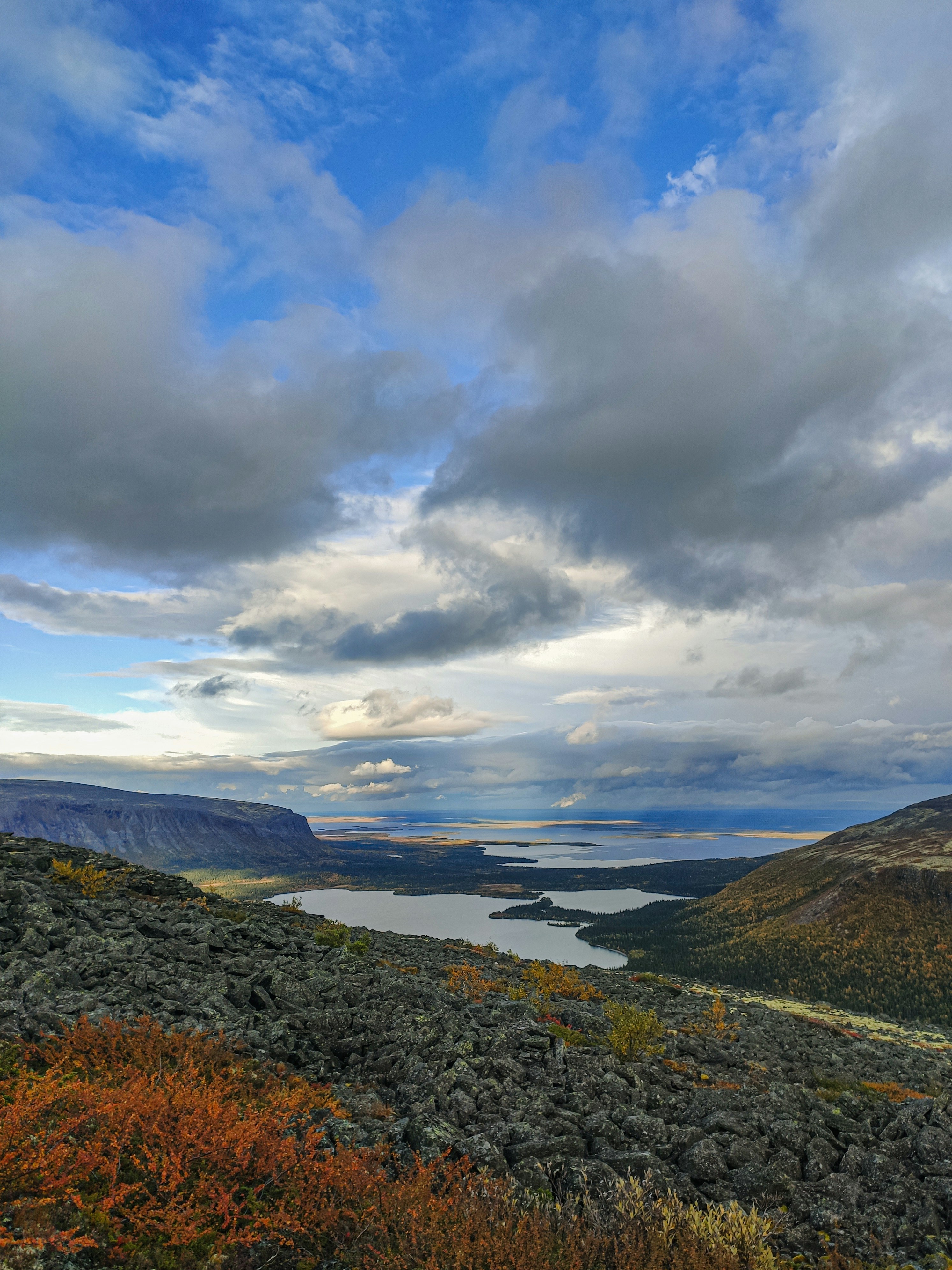 Vast landscape featuring a serene fjord surrounded by rocky terrain and colorful vegetation under a dynamic sky.