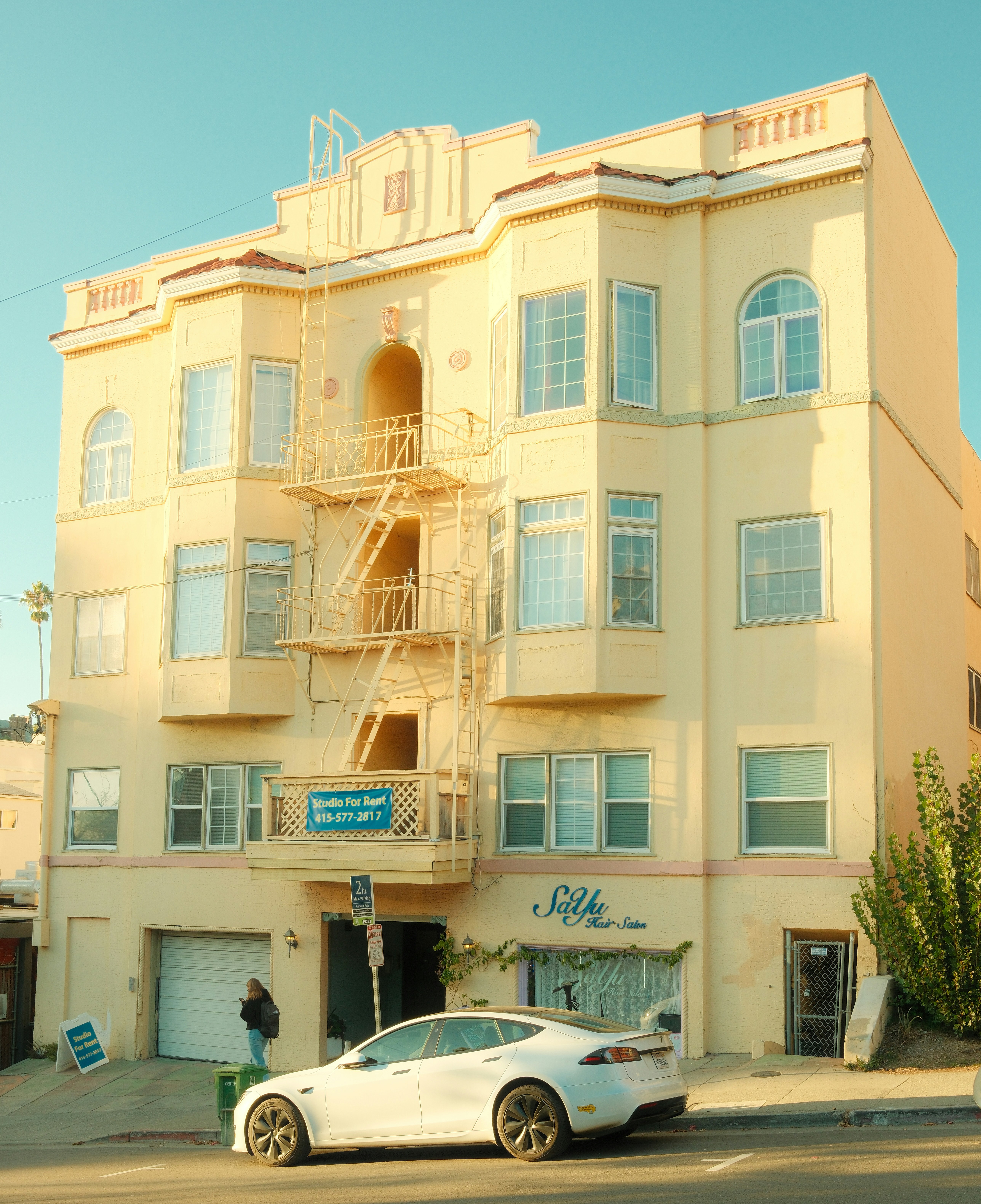 Yellow apartment building featuring a fire escape and 'For Rent' sign, surrounded by palm trees and modern vehicles.