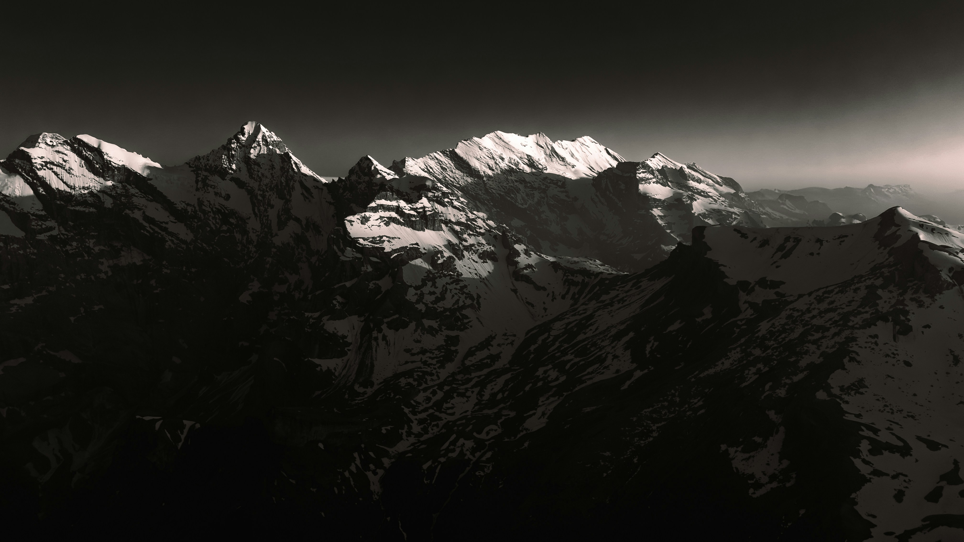 Majestic snow-capped peaks loom over a darkened landscape, capturing the essence of untouched wilderness. The interplay of light and shadow highlights the rugged terrain.