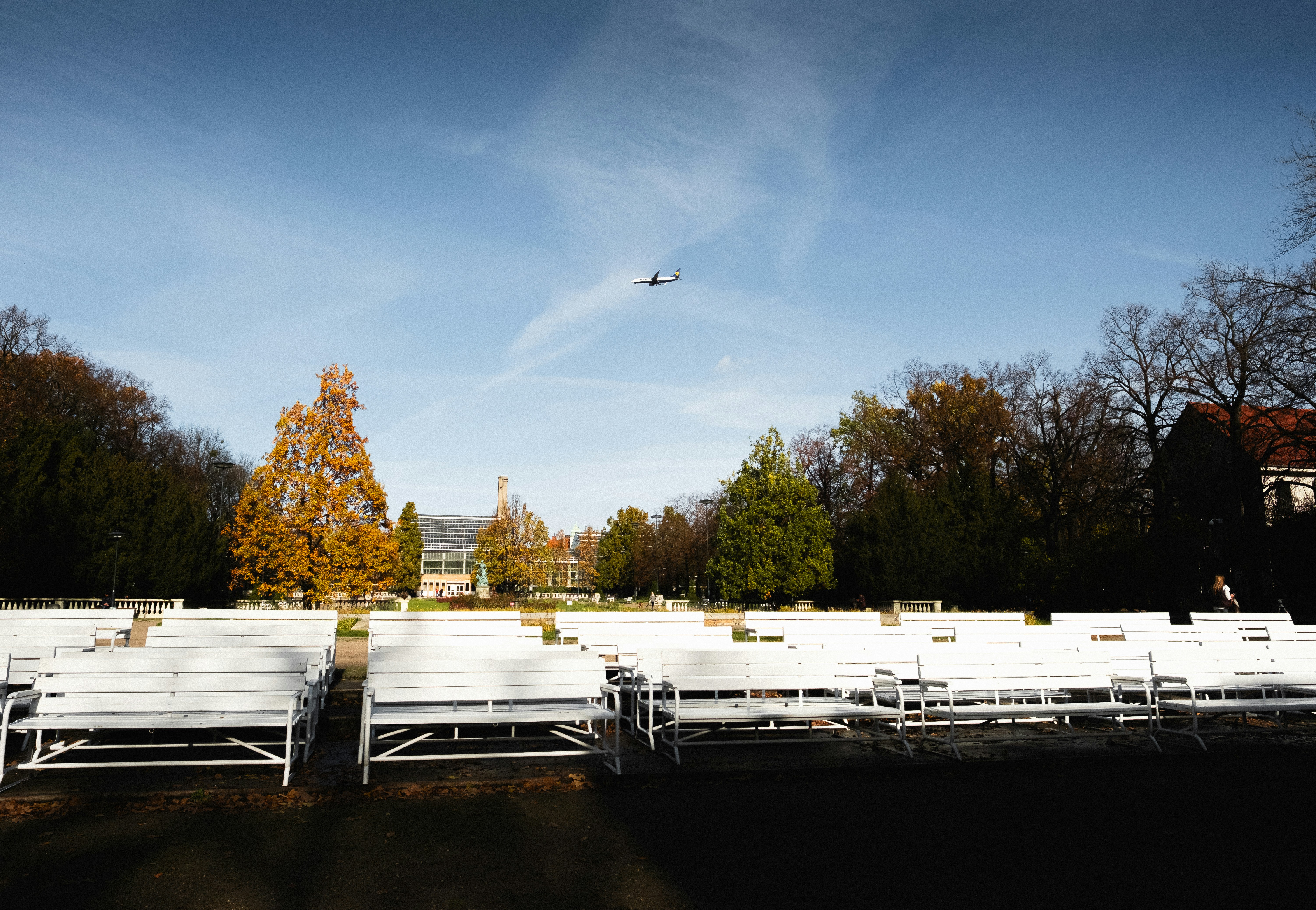 Rows of white benches in a park setting under a clear blue sky, with an airplane flying overhead and vibrant autumn trees in the background.