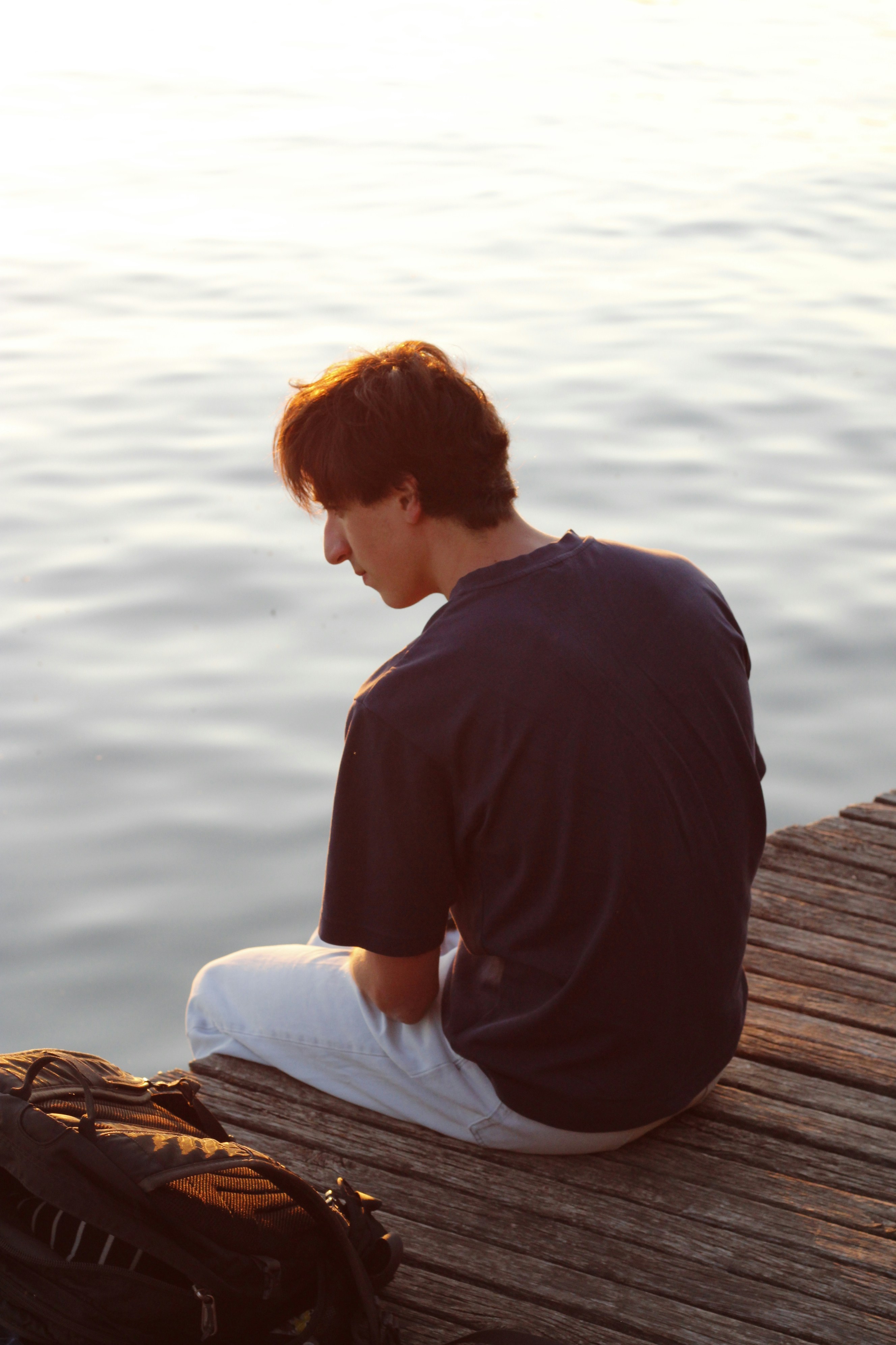 A young man sits on a wooden dock, gazing contemplatively at the water as the sun sets, casting a warm glow. A backpack rests nearby.