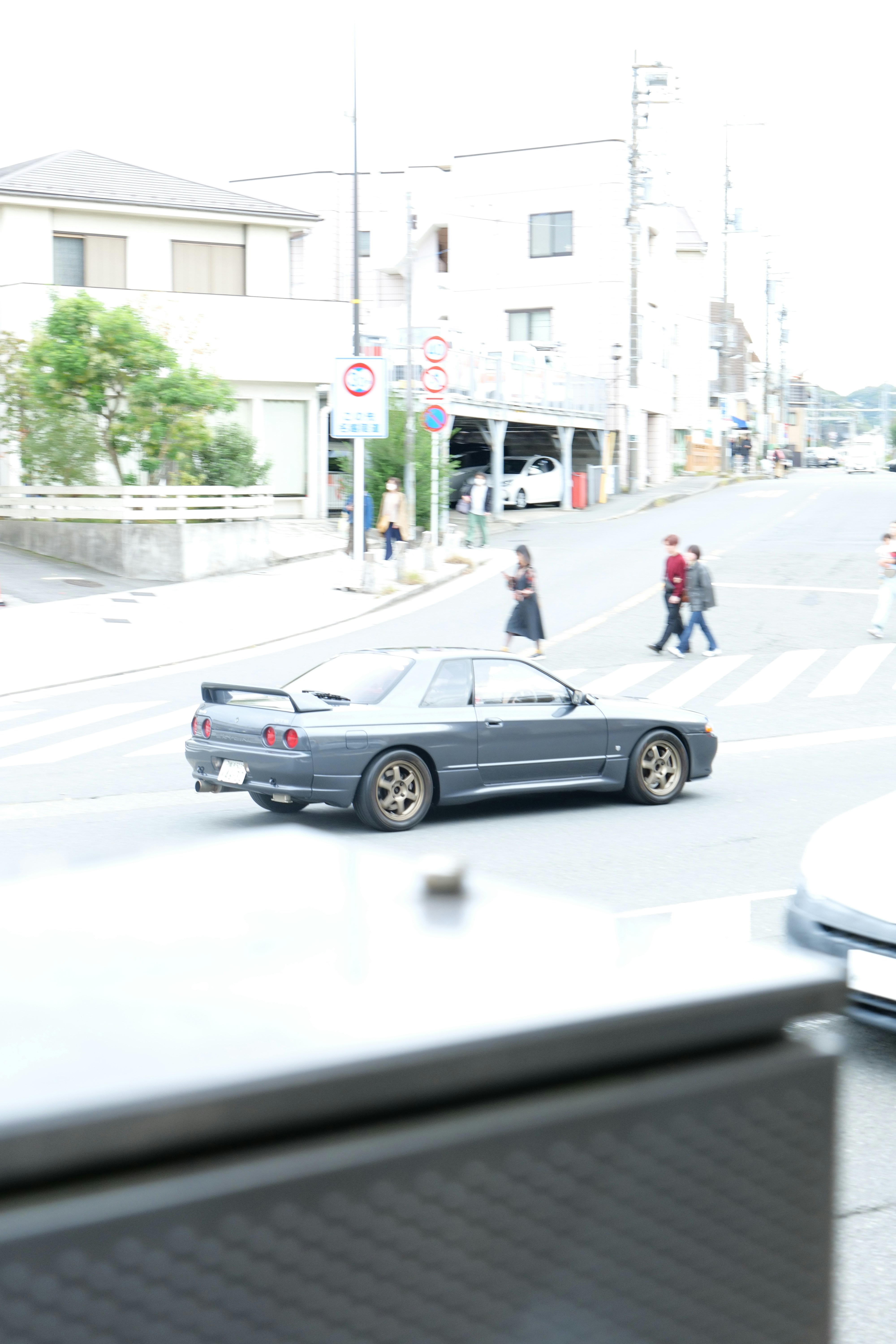 A sleek gray sports car navigates a bustling urban intersection as pedestrians cross the street. The scene captures the dynamic rhythm of city life.