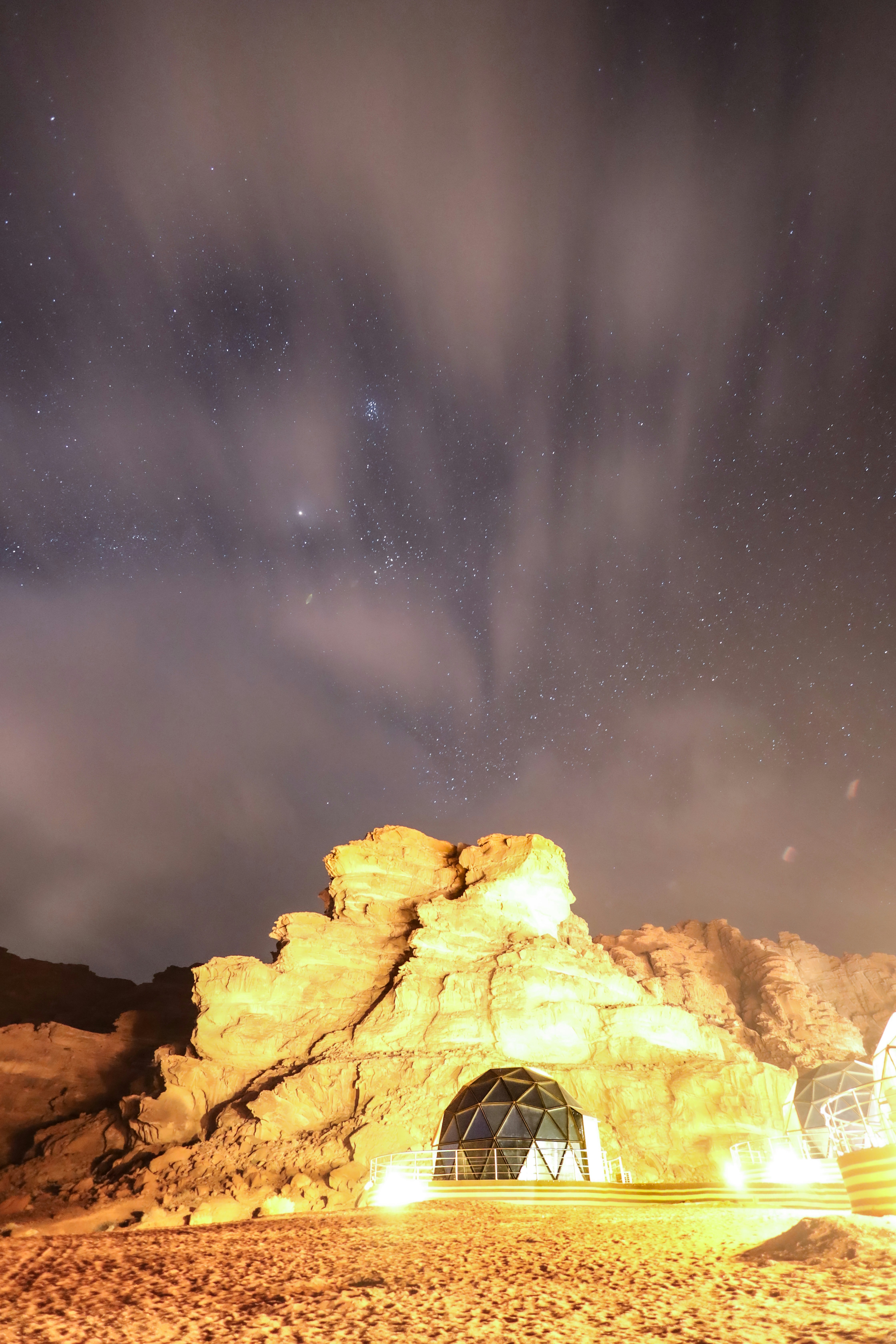 A unique rock formation illuminated by soft lights under a starry sky with wispy clouds. The scene captures the serene beauty of a desert night.